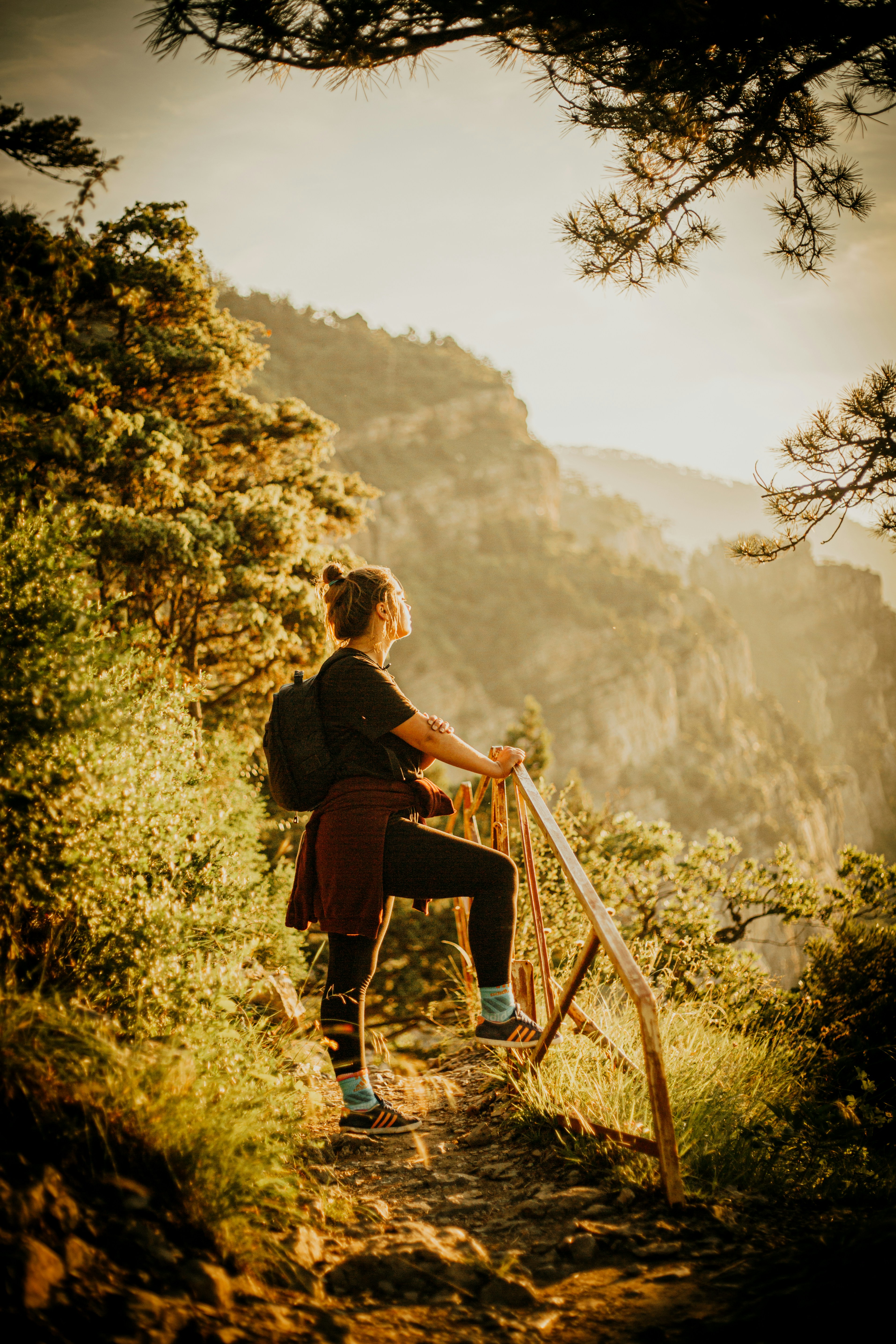 Hiker pausing on a trail, gazing out over a stunning mountainous landscape during golden hour. Sunlight filters through the trees, enhancing the serene atmosphere.