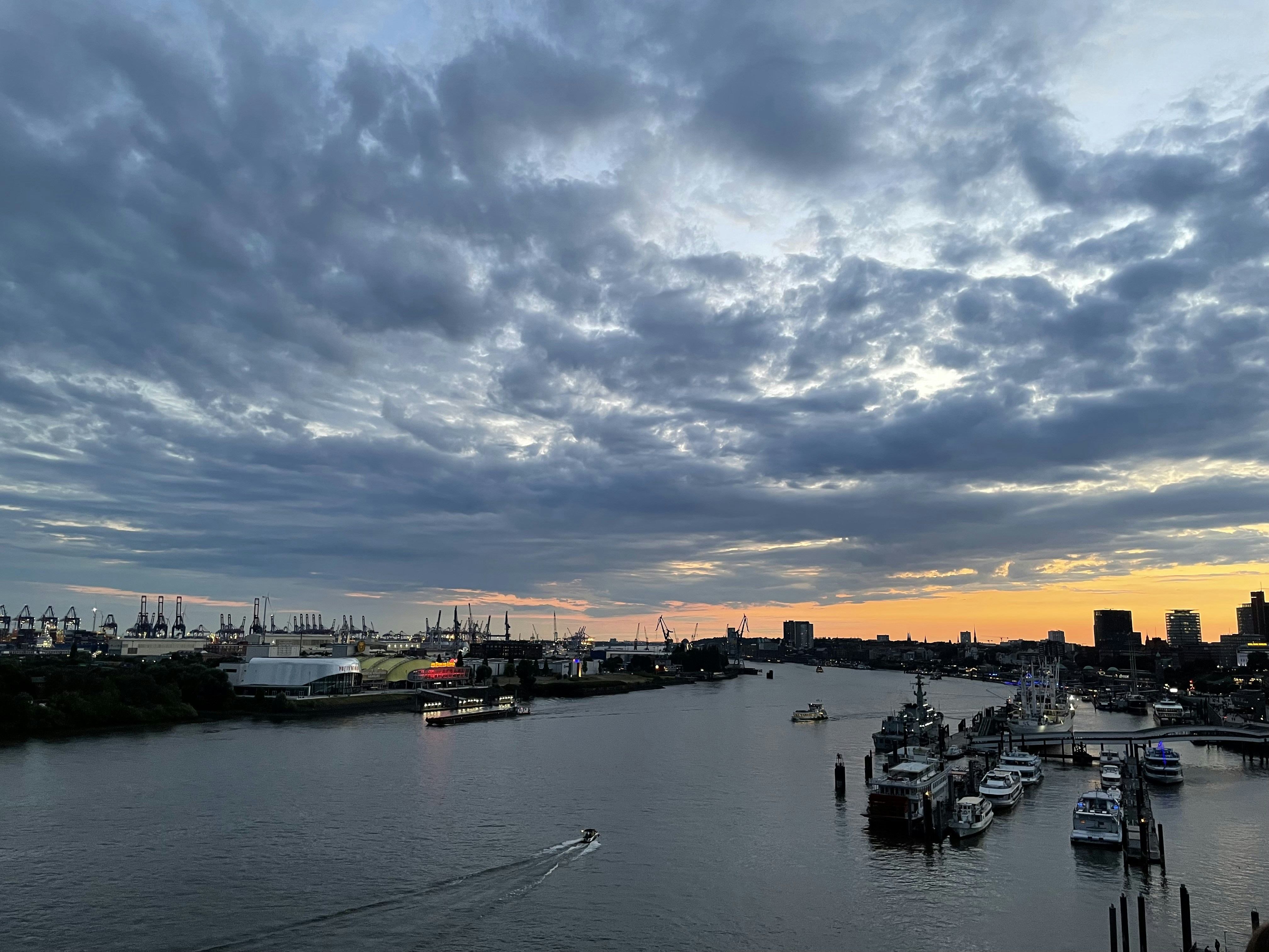 Dramatic clouds loom over a bustling river scene at dusk, with boats anchored along the shore and city lights beginning to twinkle.