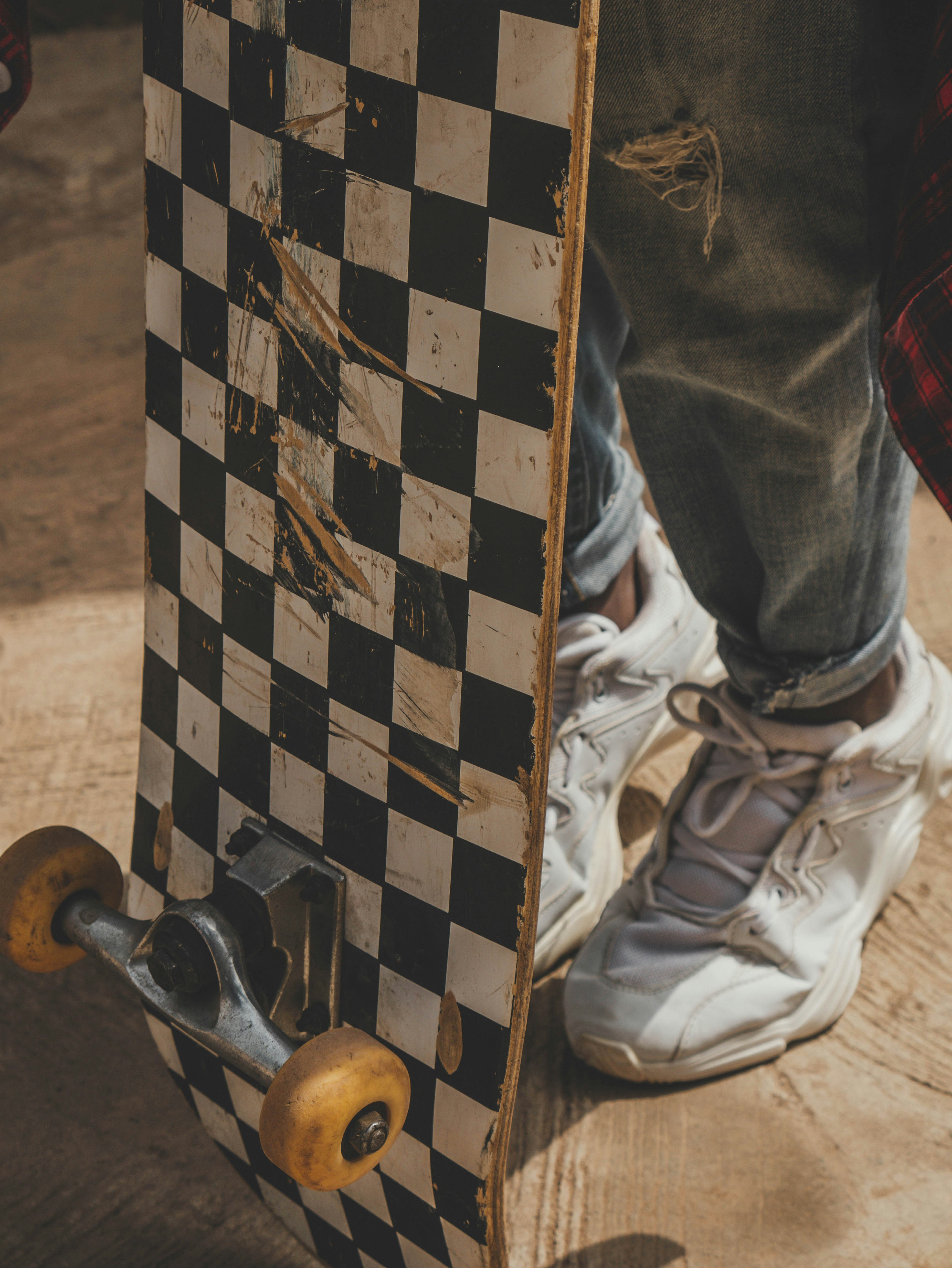 Close-up of a worn skateboard with a checkered pattern, resting beside a person's feet in casual attire. The scene captures the essence of urban skate culture.