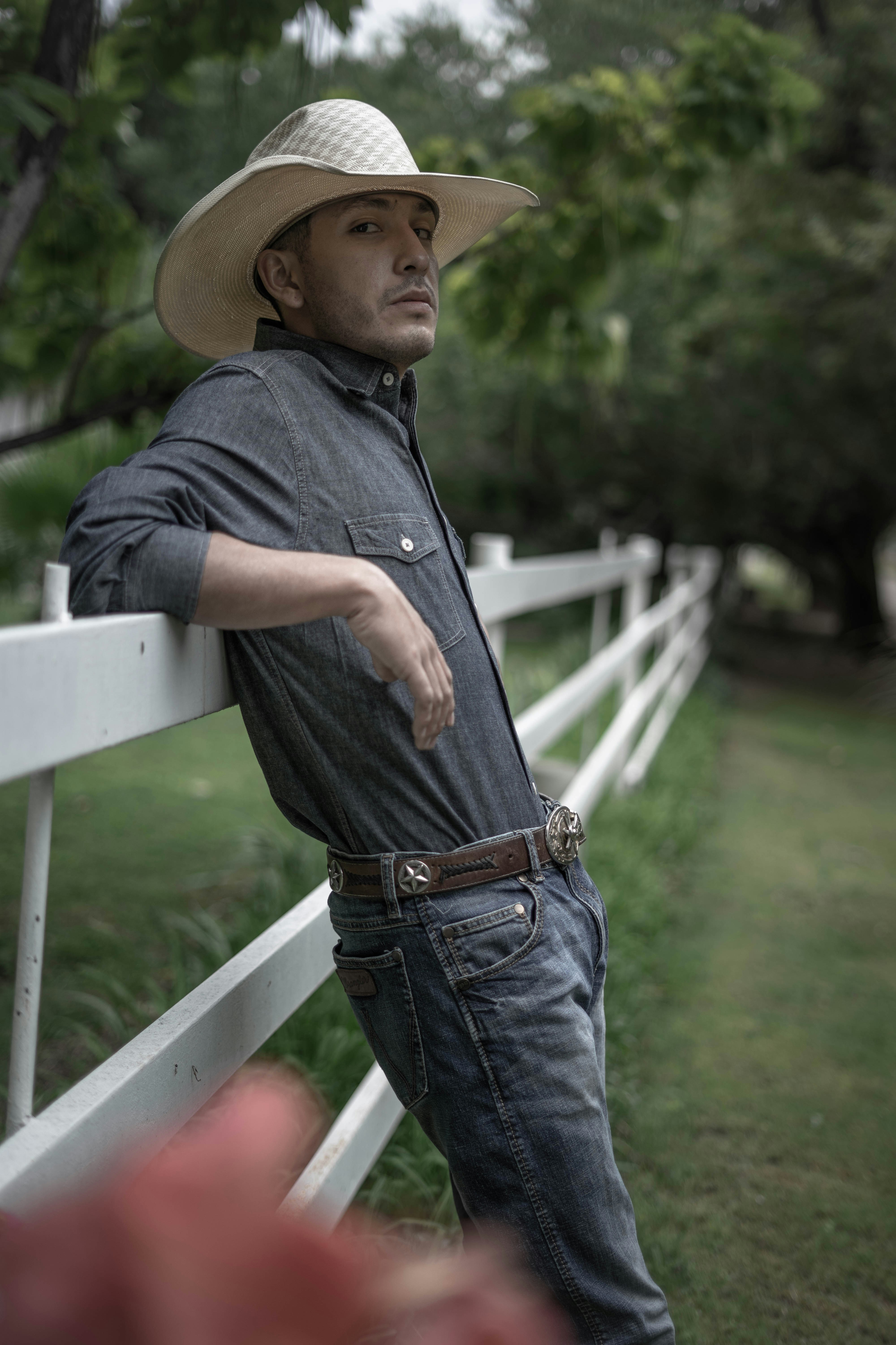 A cowboy in a denim shirt and wide-brimmed hat leans casually against a white fence, embodying a blend of tradition and contemporary style.