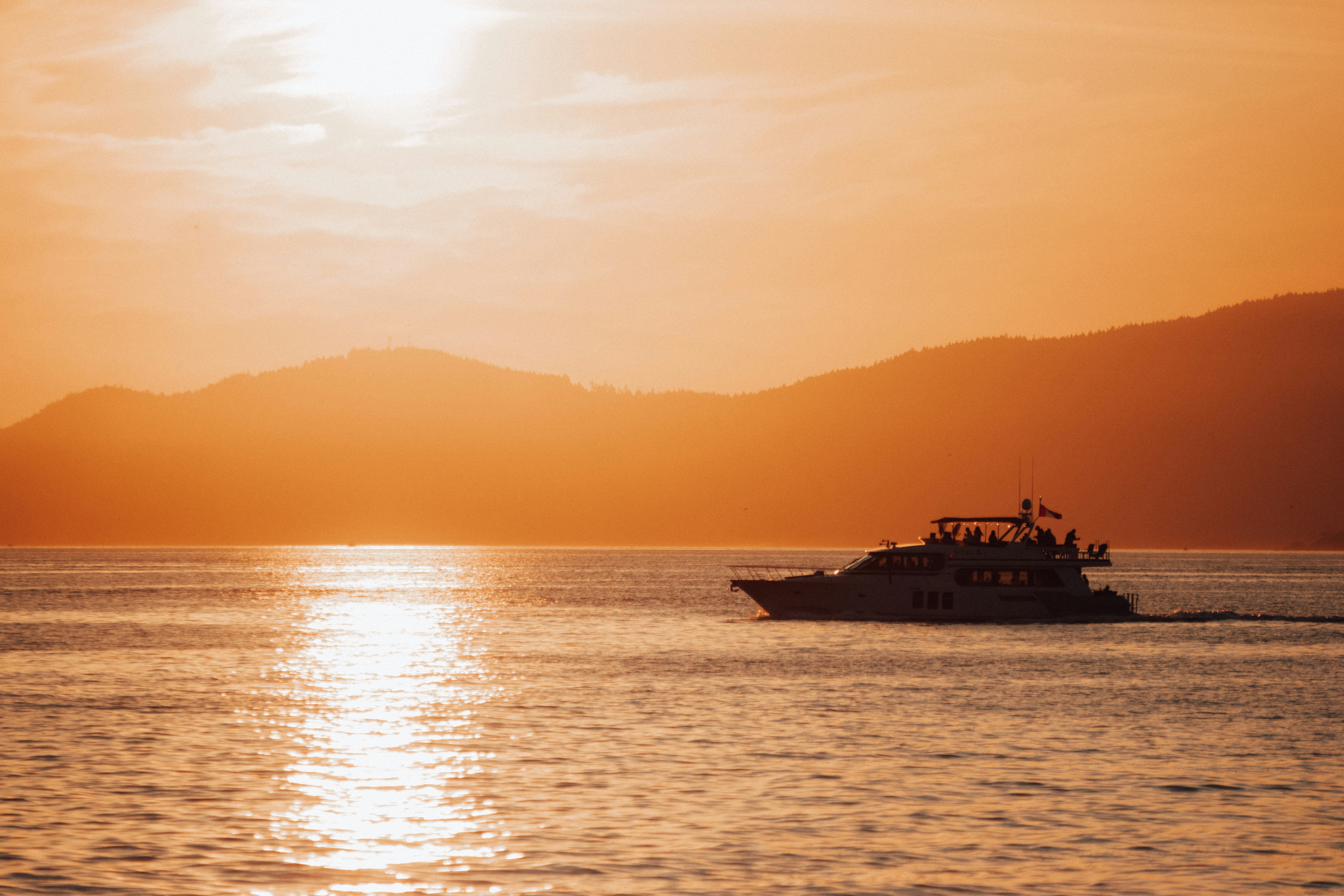 Silhouette of a ship gliding across a sunlit sea with distant mountains under a glowing sunset.