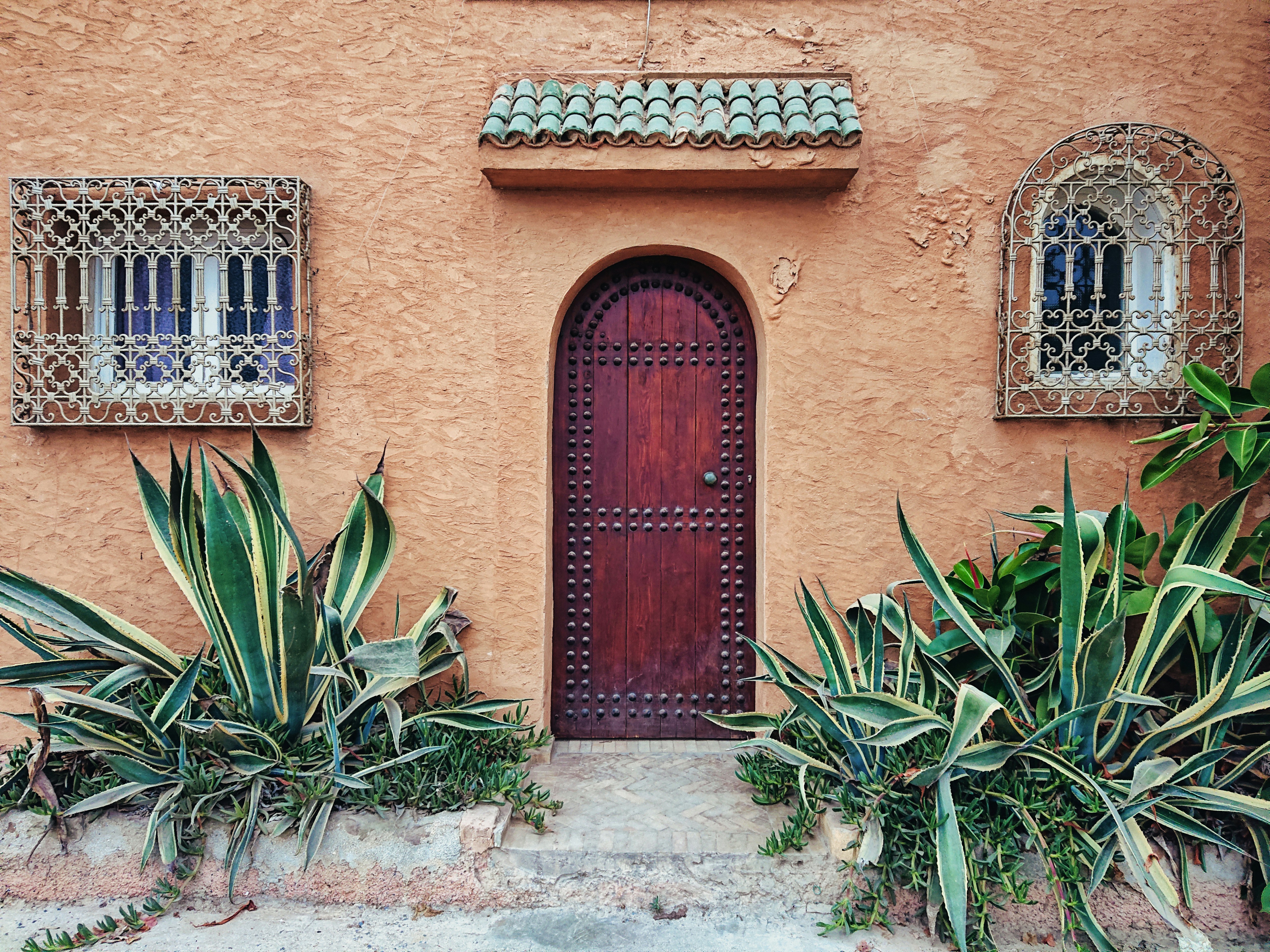 Terracotta doorway with studded wooden door, flanked by decorative iron windows, surrounded by spiky agave plants.