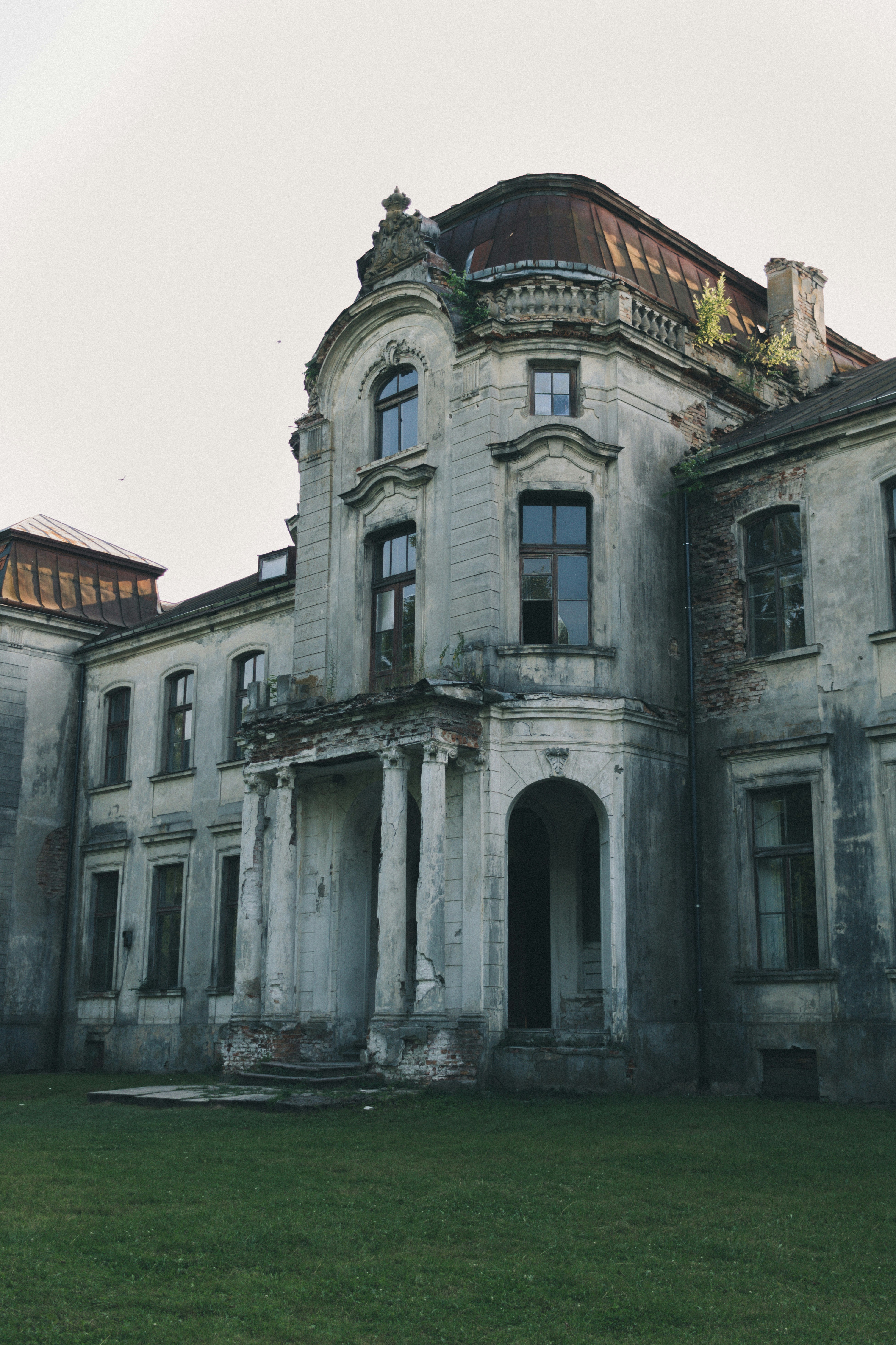 An abandoned mansion showcasing intricate architectural details, with crumbling columns and a weathered façade surrounded by overgrown grass.