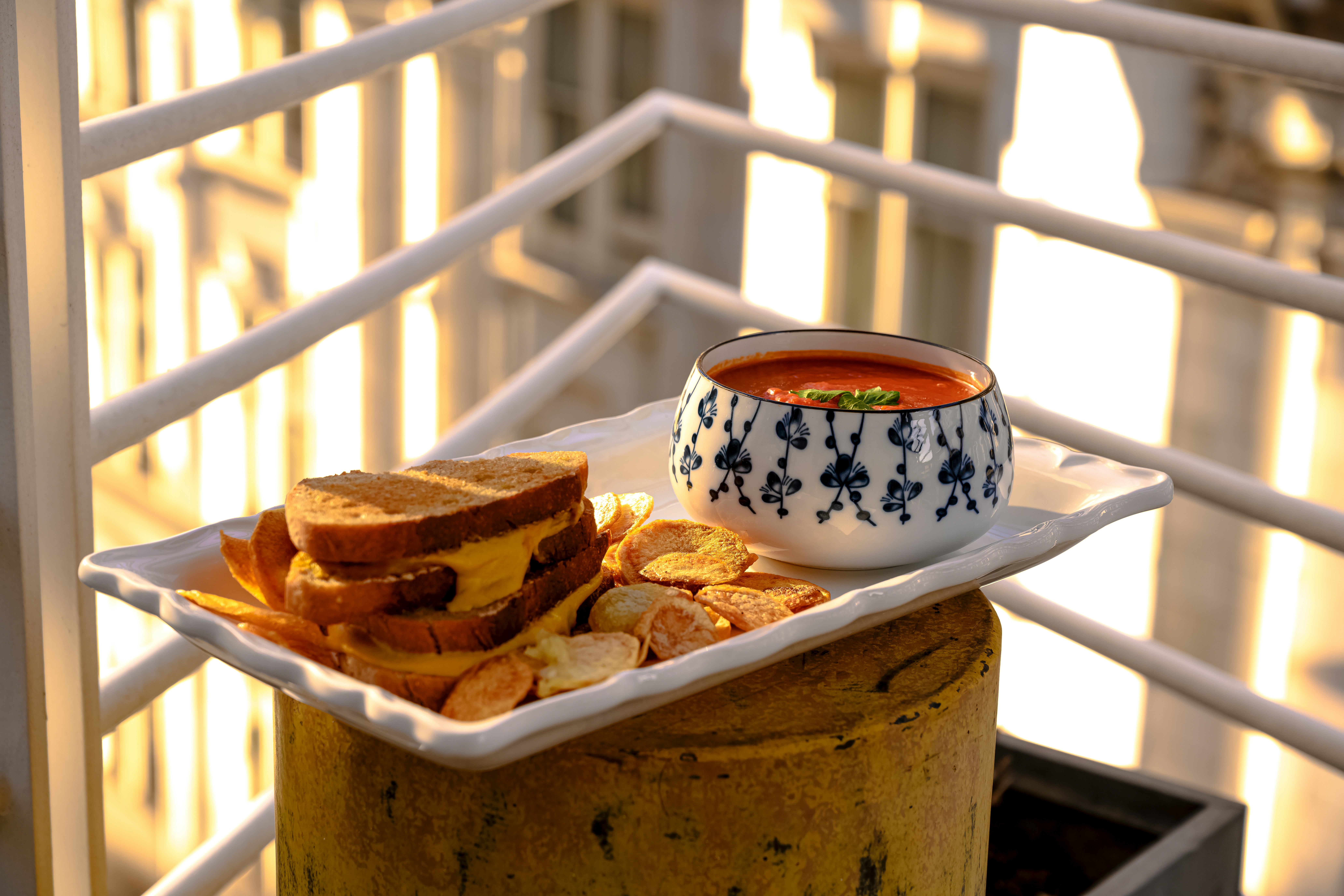 Grilled cheese sandwich and crispy chips served with a bowl of tomato soup, set against a sunlit balcony backdrop.
