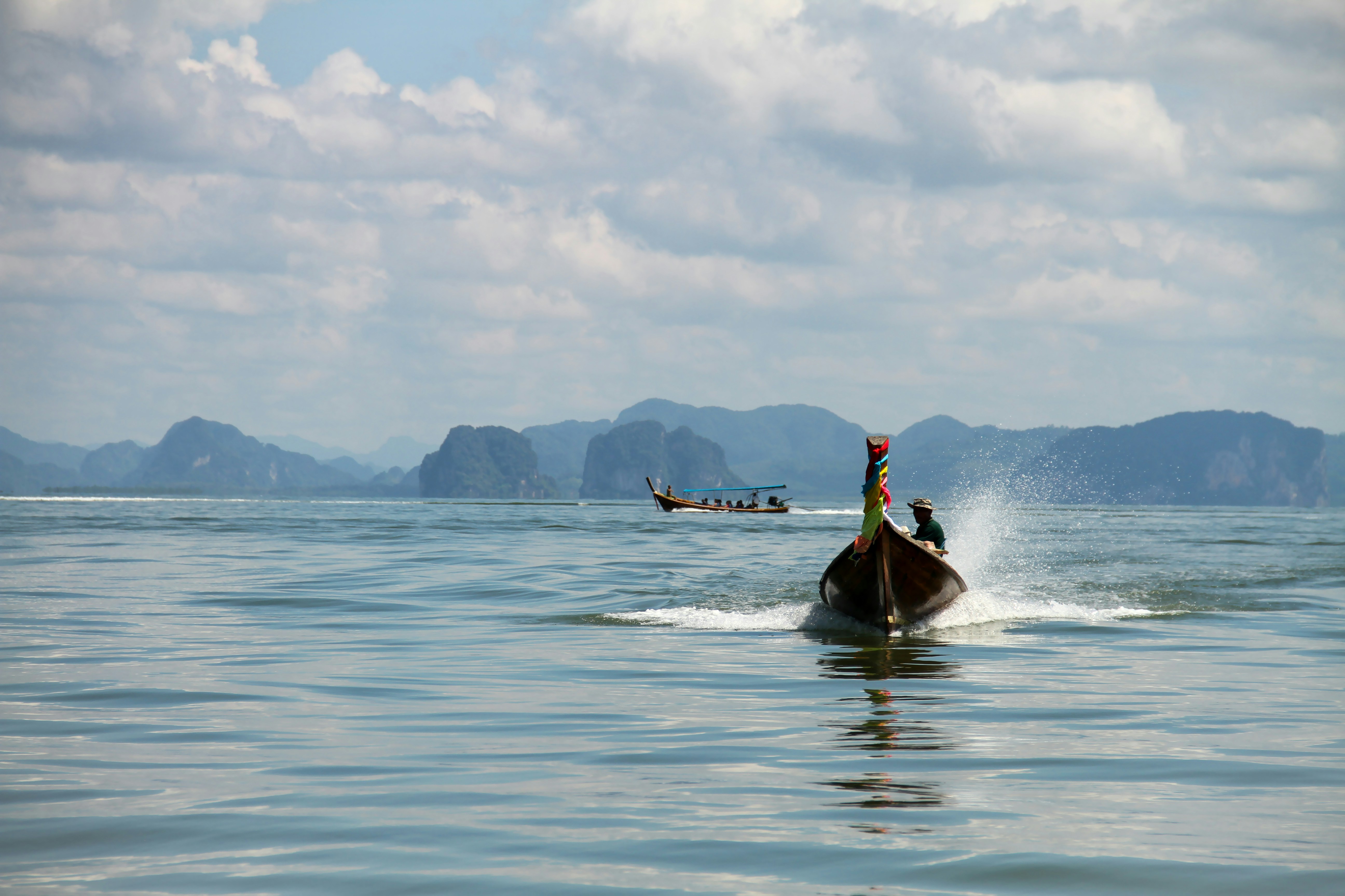 Longtail boat speeding across calm waters, with distant limestone cliffs and a second boat in the background. 