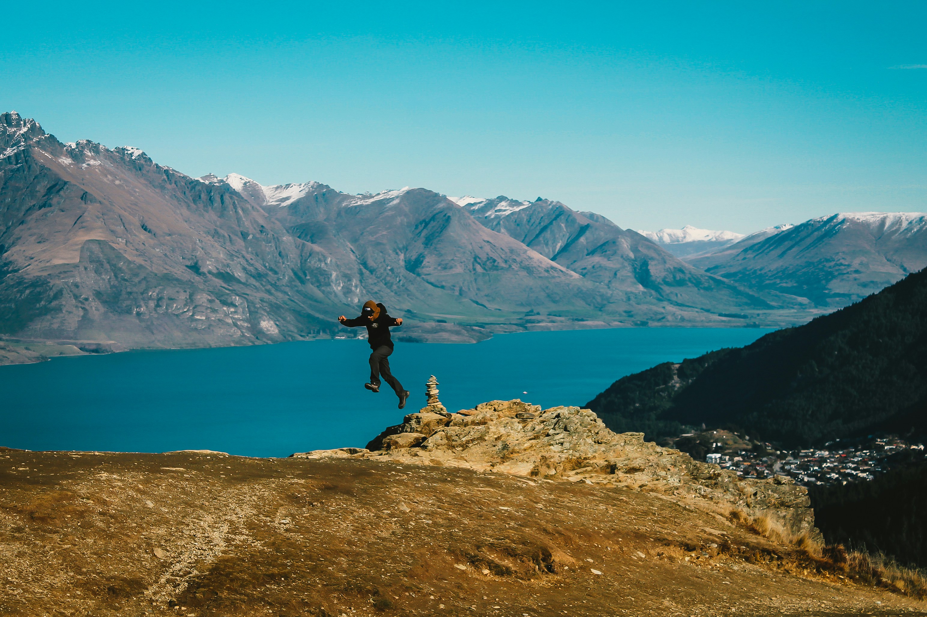This captivating image captures a person mid-jump on a rugged mountain ledge, set against a breathtaking backdrop of expansive blue waters and towering peaks. The vibrant hues of the clear sky and the deep blue lake contrast beautifully with the earthy tones of the rocky terrain, creating a striking visual composition. The bright daylight enhances the crispness and clarity, conveying a sense of adventure and exhilaration.