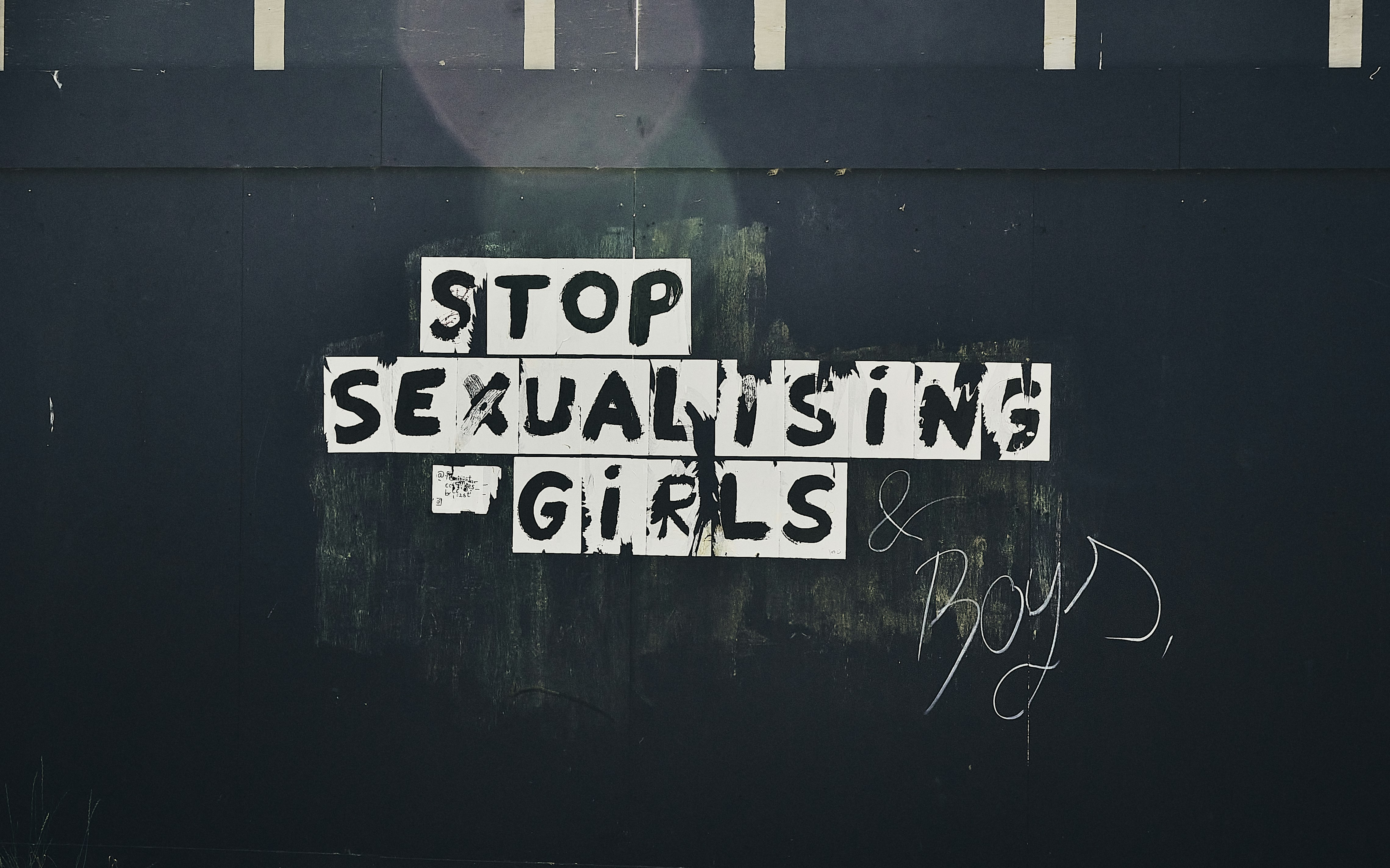 A bold street art message urging to stop the sexualization of girls, displayed on a textured dark wall with graffiti elements.