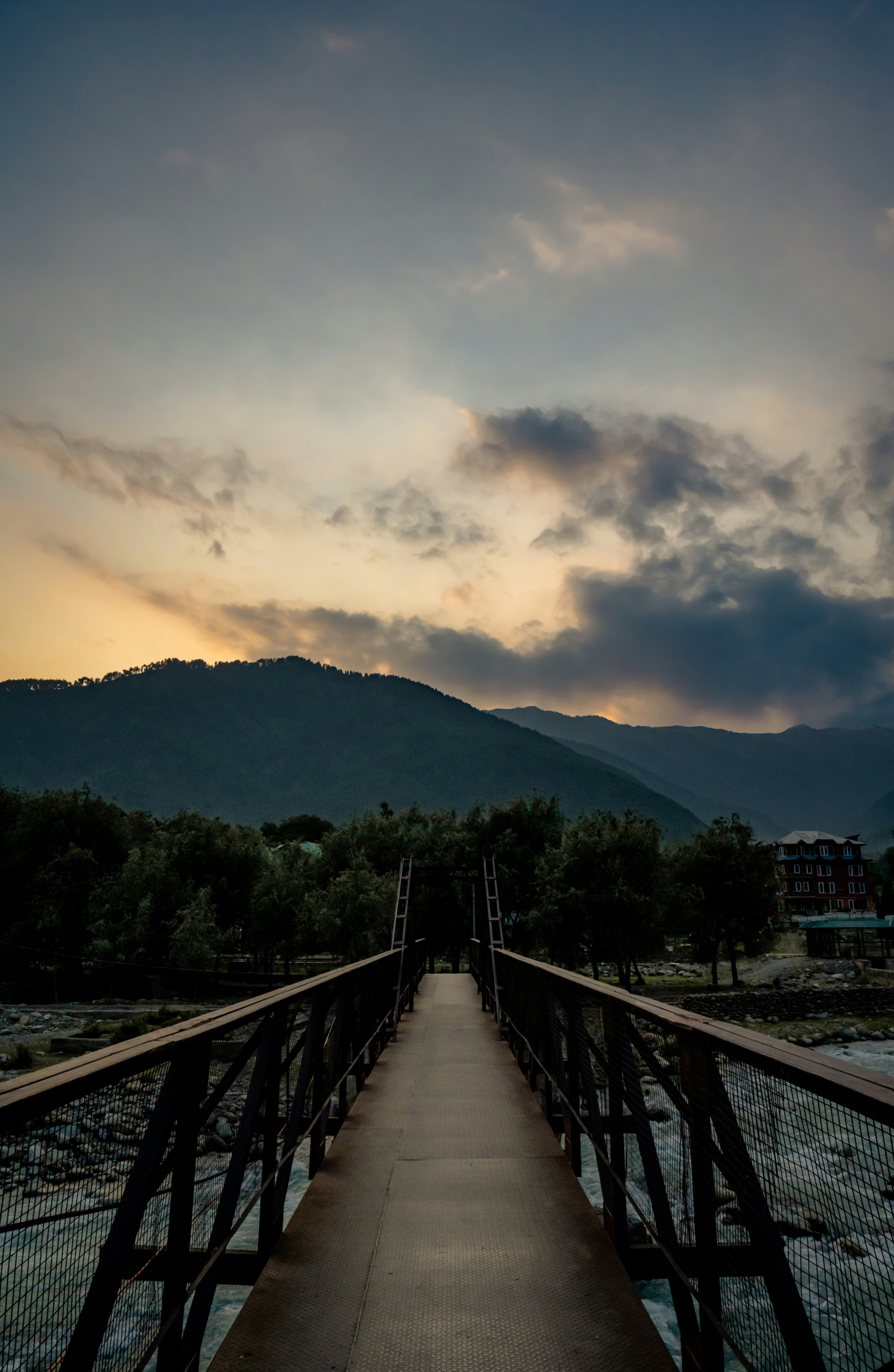 Rustic metal bridge leading towards distant mountains under a twilight sky. The scene evokes a sense of adventure and calm.
