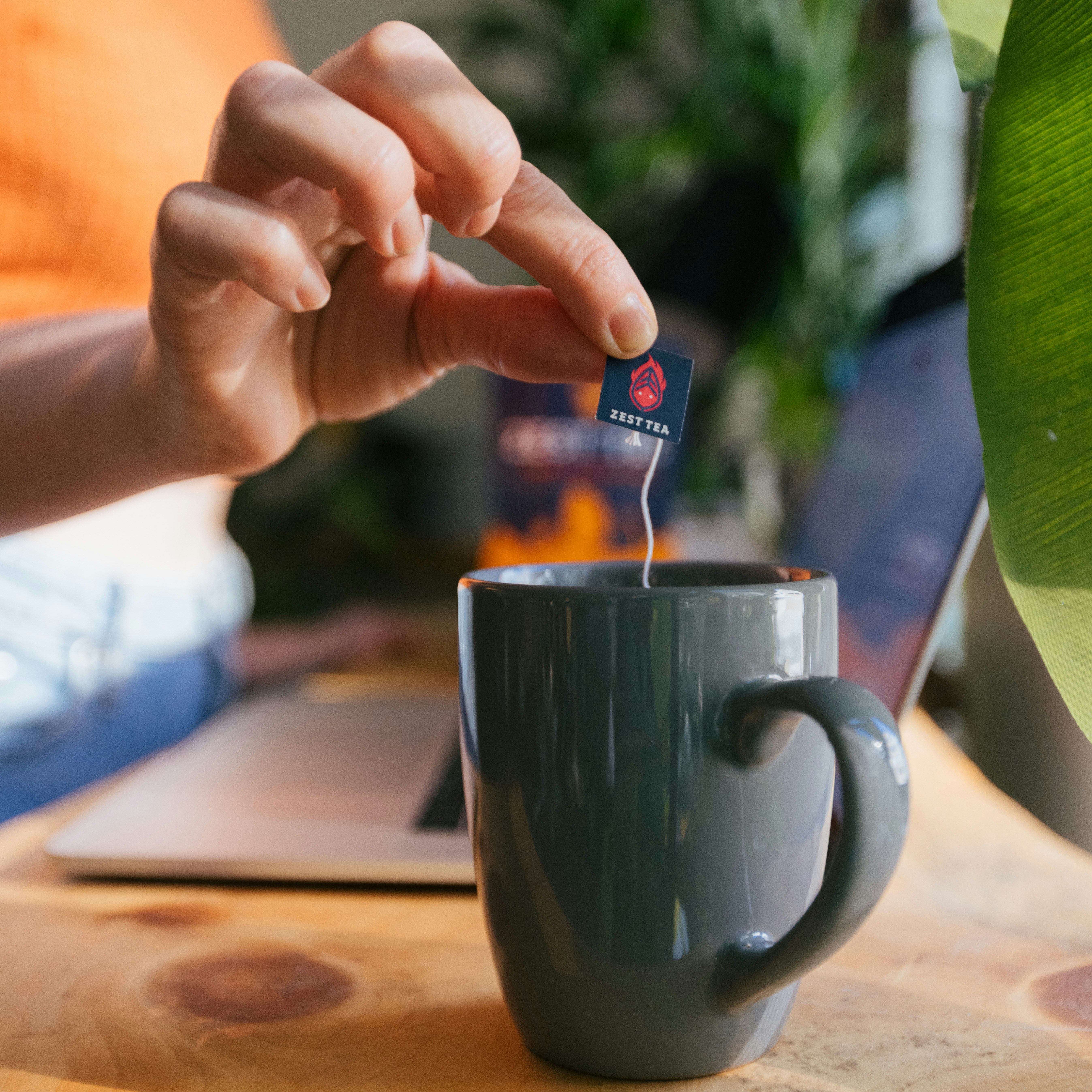 Hand placing a tea bag into a gray mug on a wooden table, with a laptop and a vibrant tea box in the background.