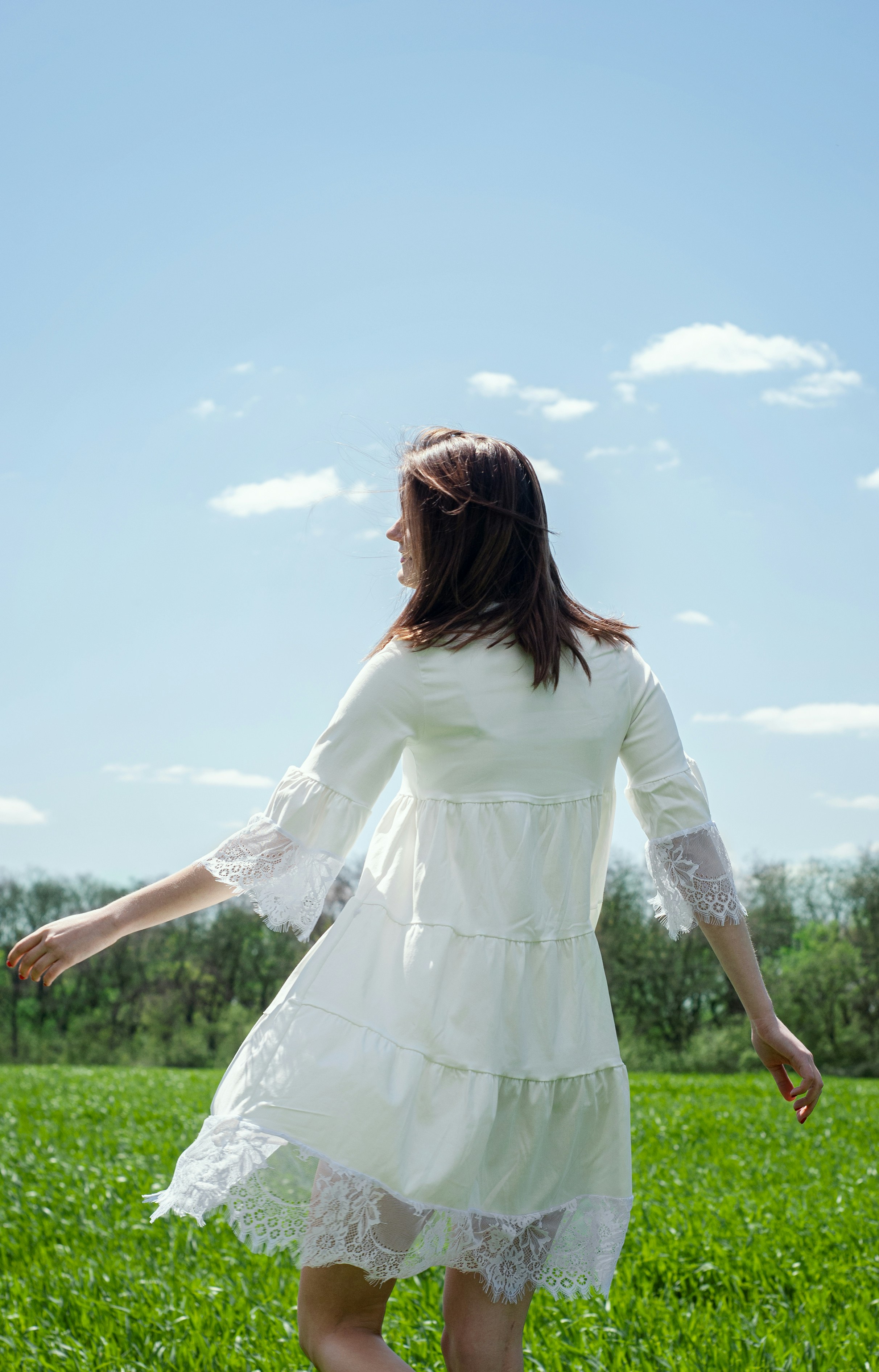 A woman twirls gracefully in a white lace dress amidst a vibrant green field under a bright blue sky. The scene conveys a sense of freedom and connection with nature.