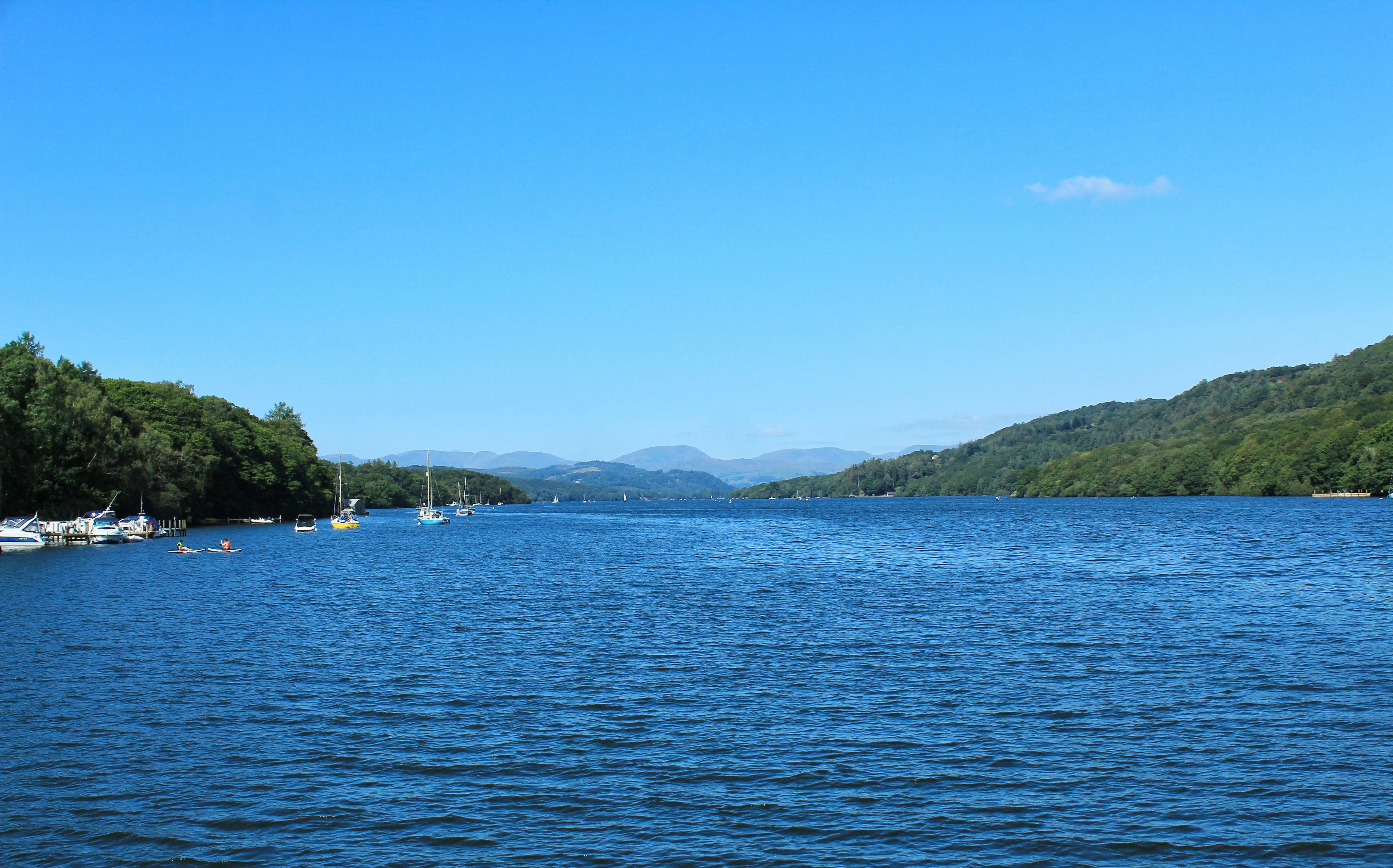 Tranquil lake scene with boats moored along the shore, surrounded by lush greenery and distant hills under a clear blue sky.