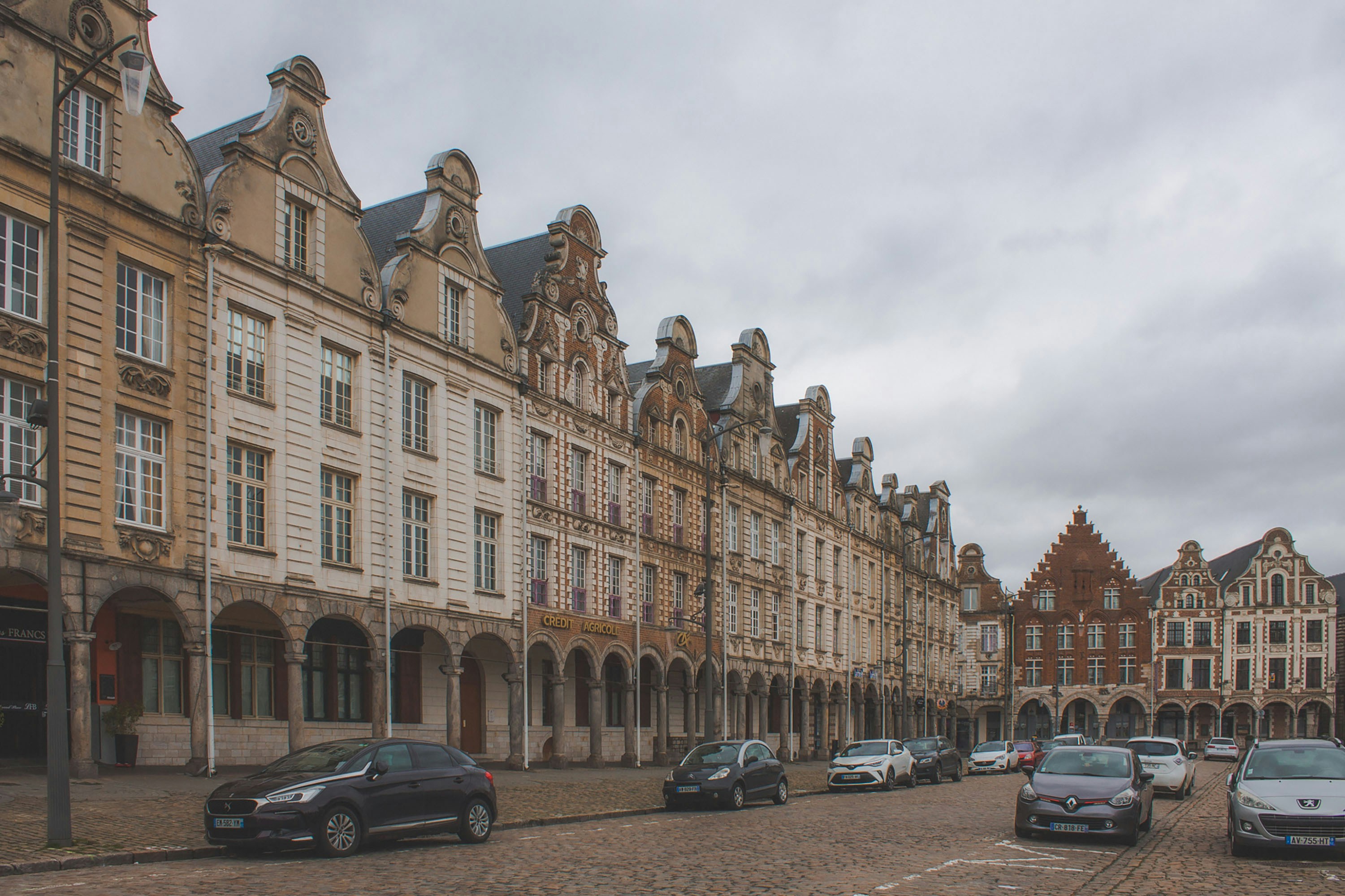 Row of historic European buildings with arched walkways and parked cars under a cloudy sky.
