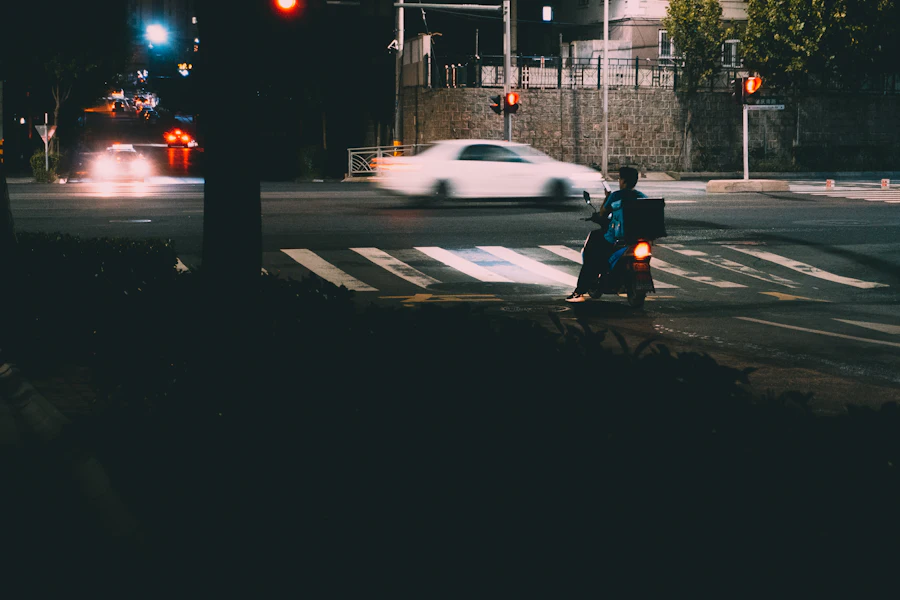 A scooter rider waits at a dimly lit intersection as a white car blurs past and traffic lights glow above. Dark foreground shapes frame the nighttime street scene.