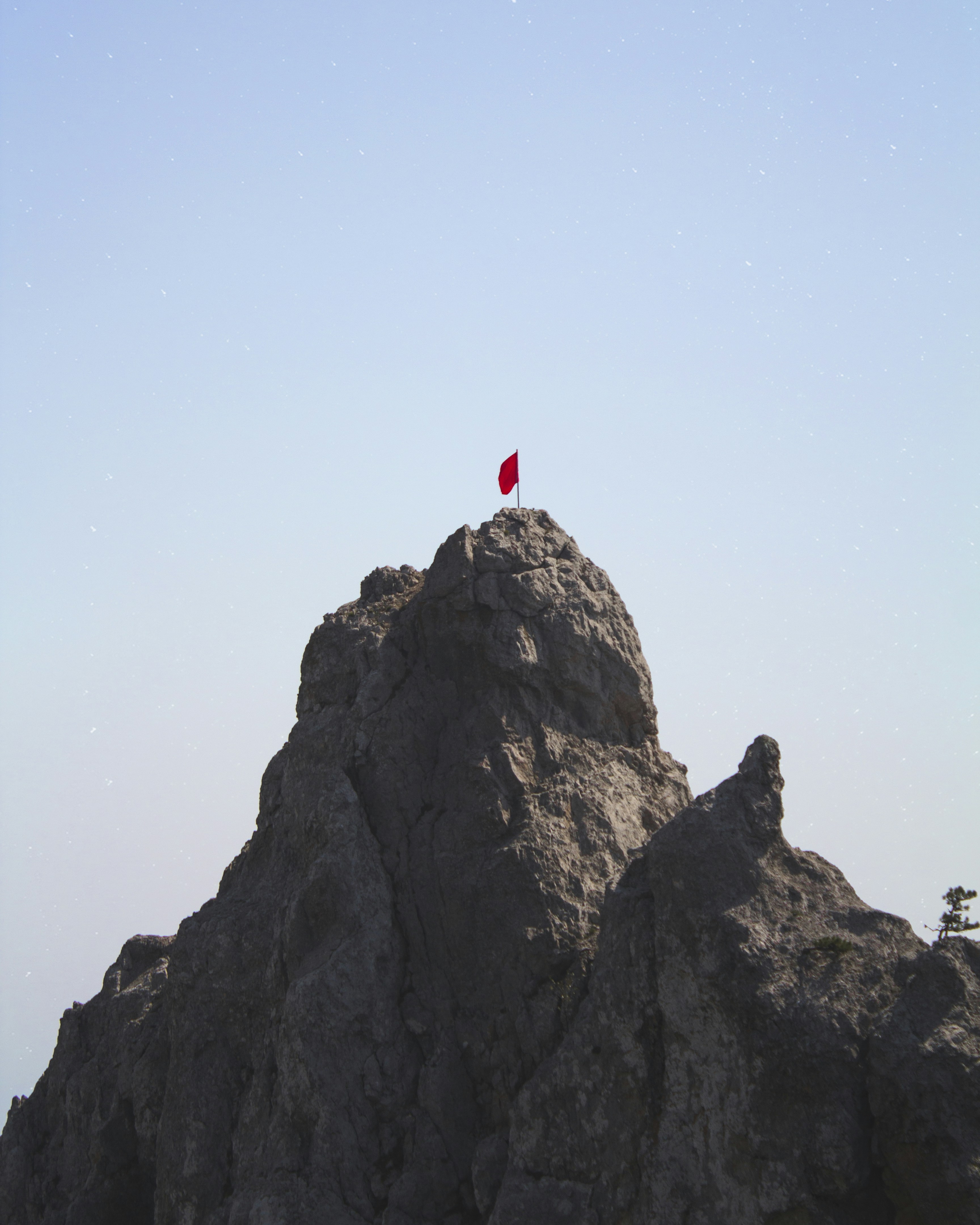 A rugged mountain peak crowned with a red flag against a clear sky, symbolizing triumph and perseverance.