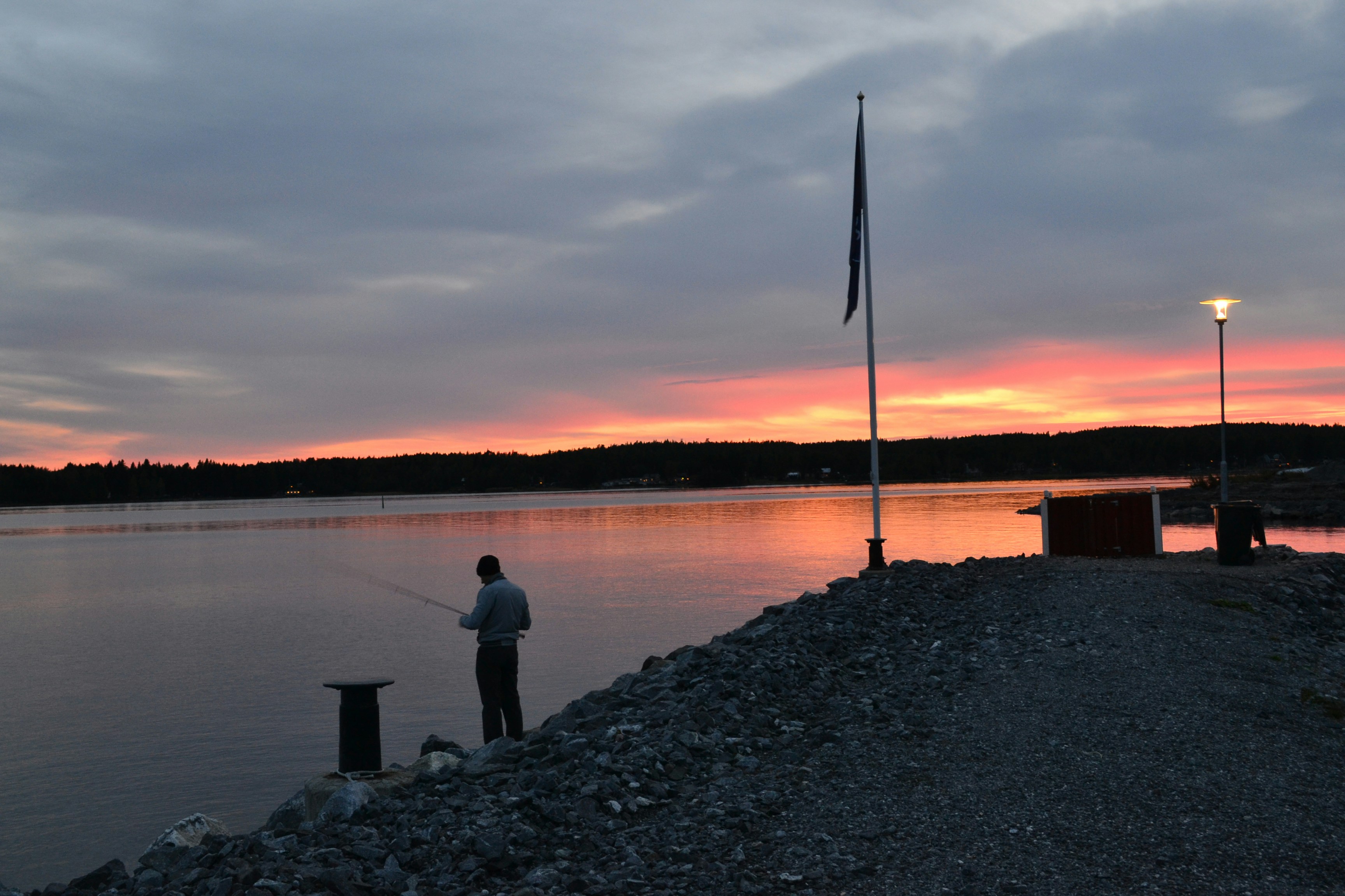 Fisherman casting a line into a tranquil lake at sunset, with vibrant colors reflecting on the water's surface.