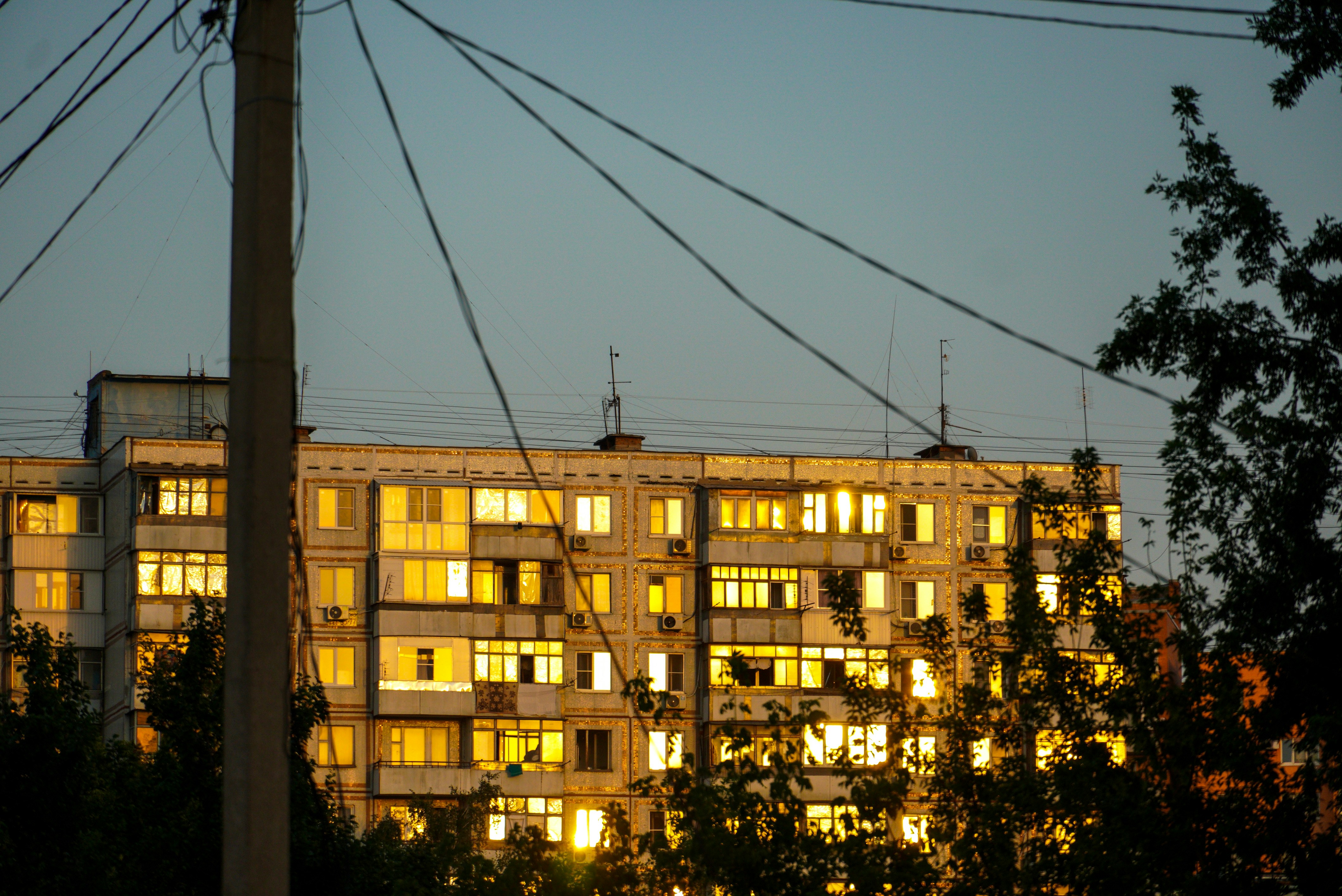 Illuminated windows of a residential building create a warm glow against the evening sky, framed by silhouetted trees and power lines.