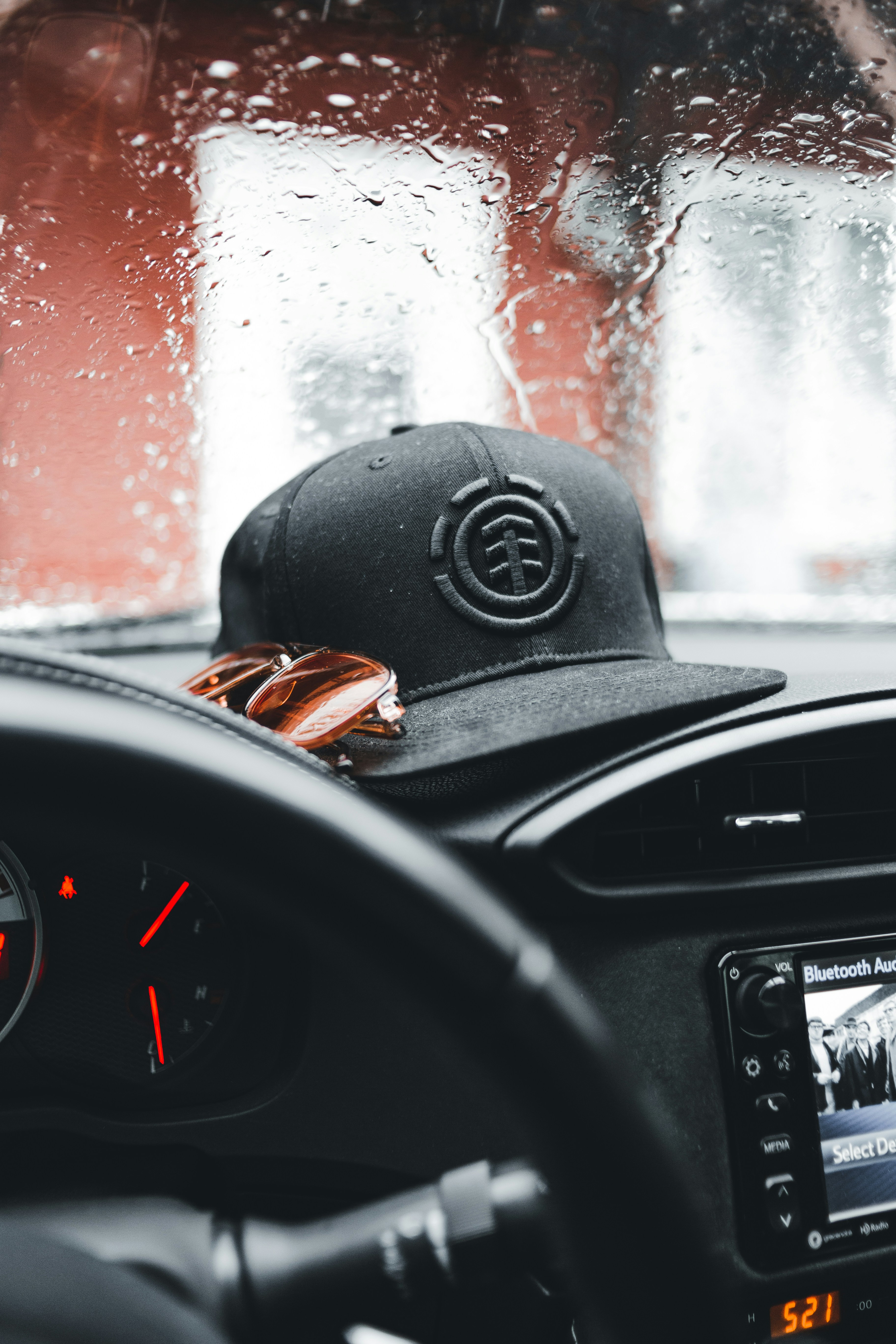 A black cap rests on the dashboard of a car, accompanied by sunglasses, with raindrops adorning the windshield, hinting at a rainy day.