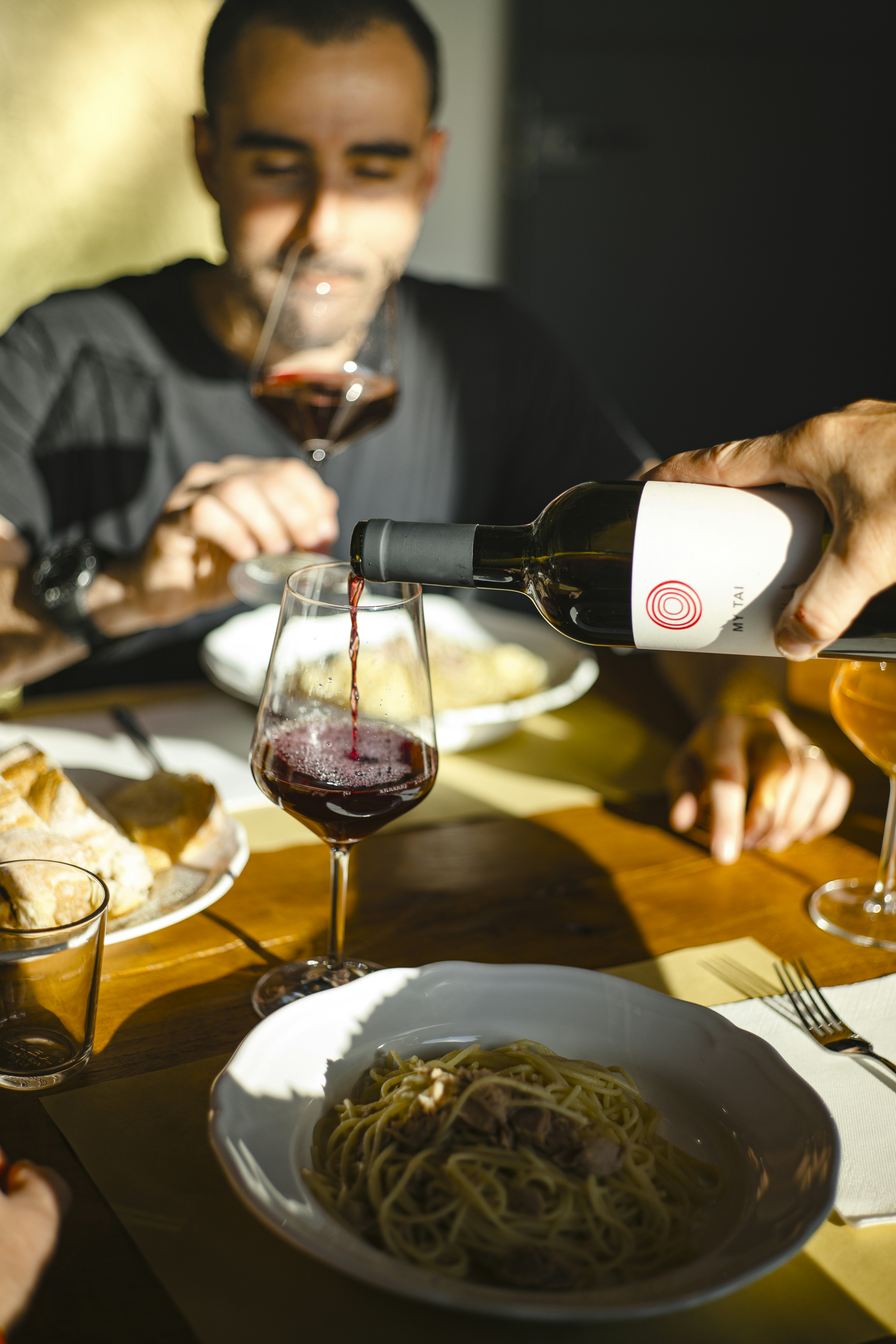 A hand pours red wine into a glass while a man enjoys a meal of pasta at a dining table, surrounded by bread and other dishes.