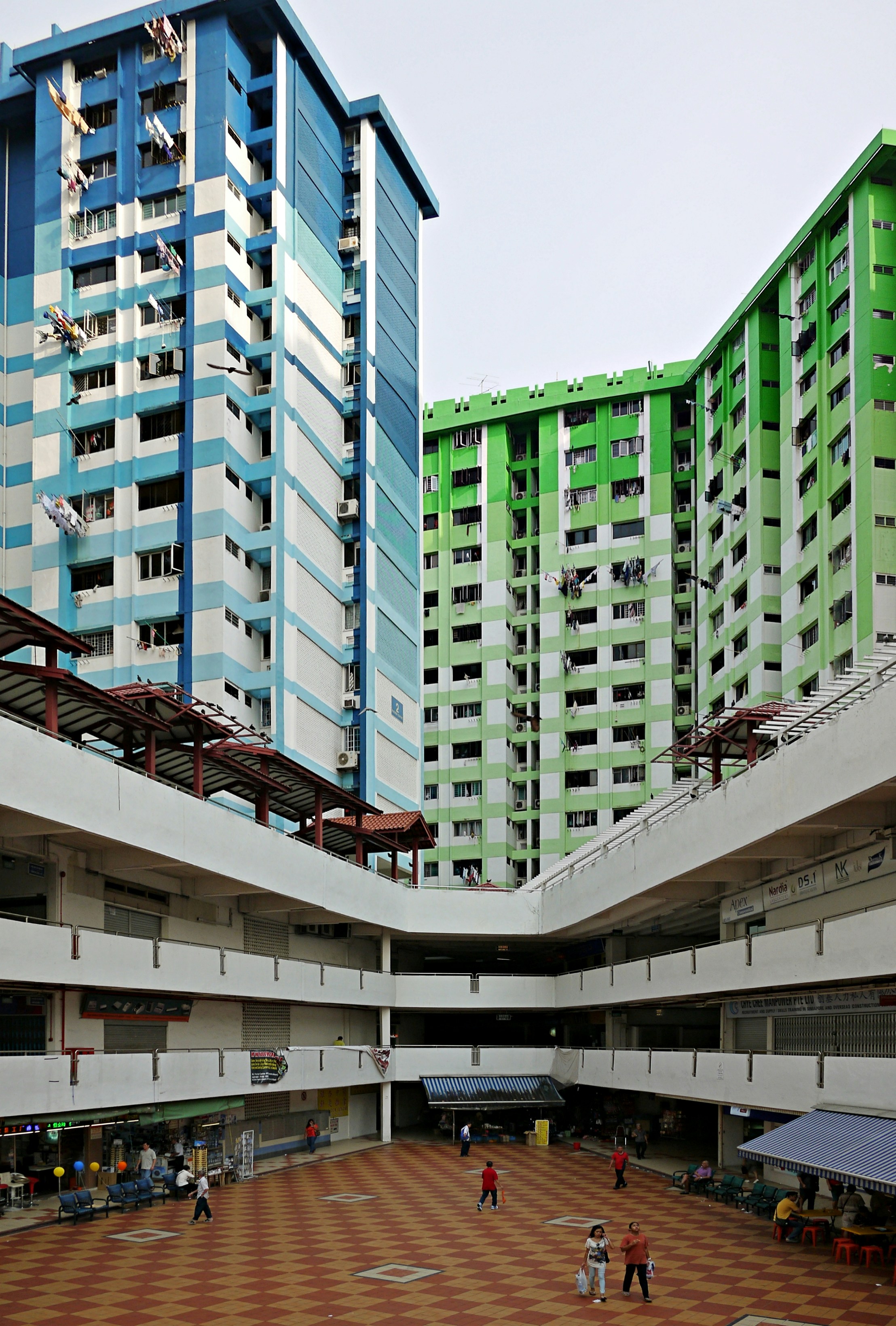 Photograph of a colorful apartment complex with blue and green towers framing a tiled courtyard. People stroll through the open plaza, highlighting urban geometry and everyday life.