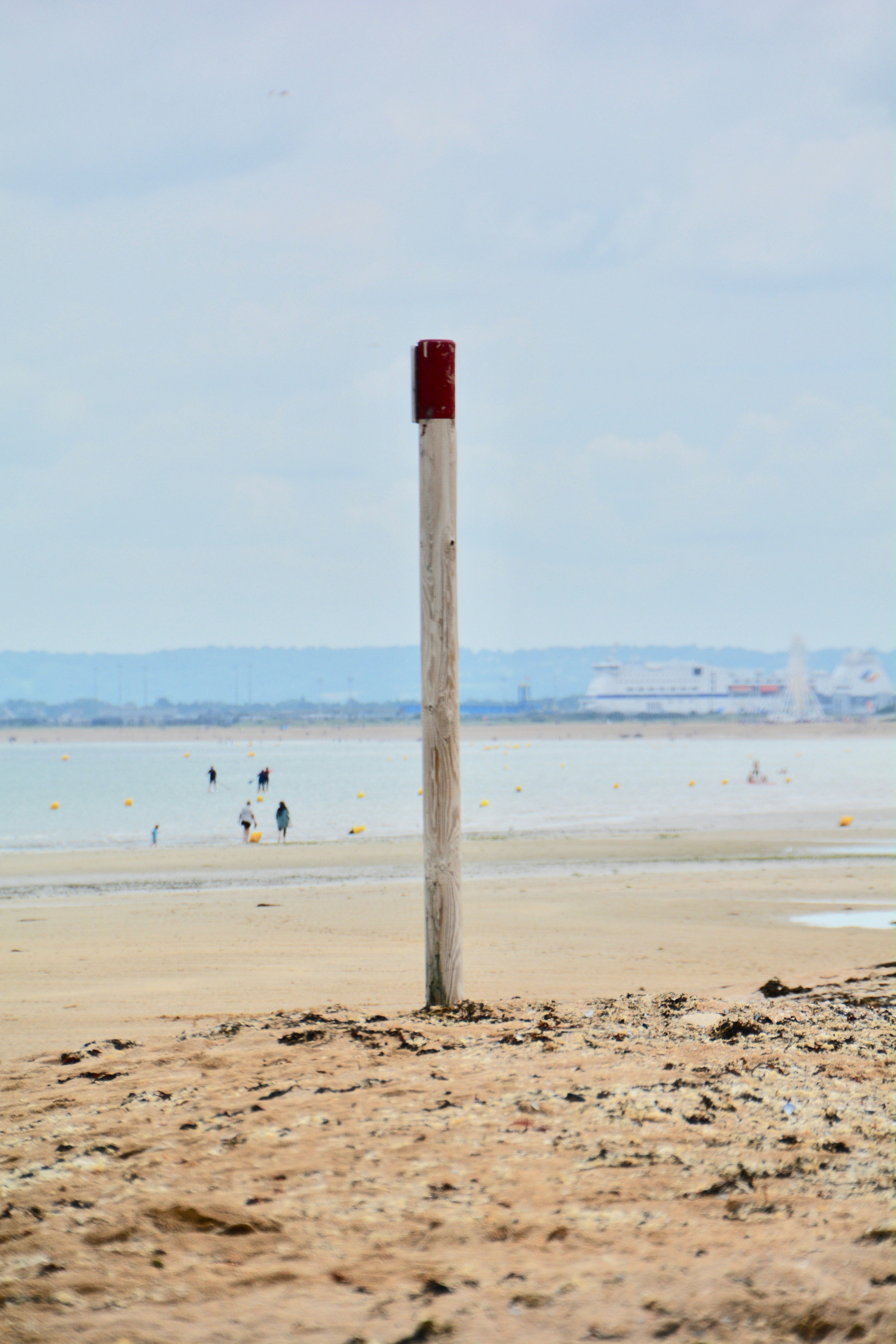 A weathered pole with a red top stands on a sandy beach, overlooking a calm sea where people stroll and enjoy the shoreline.