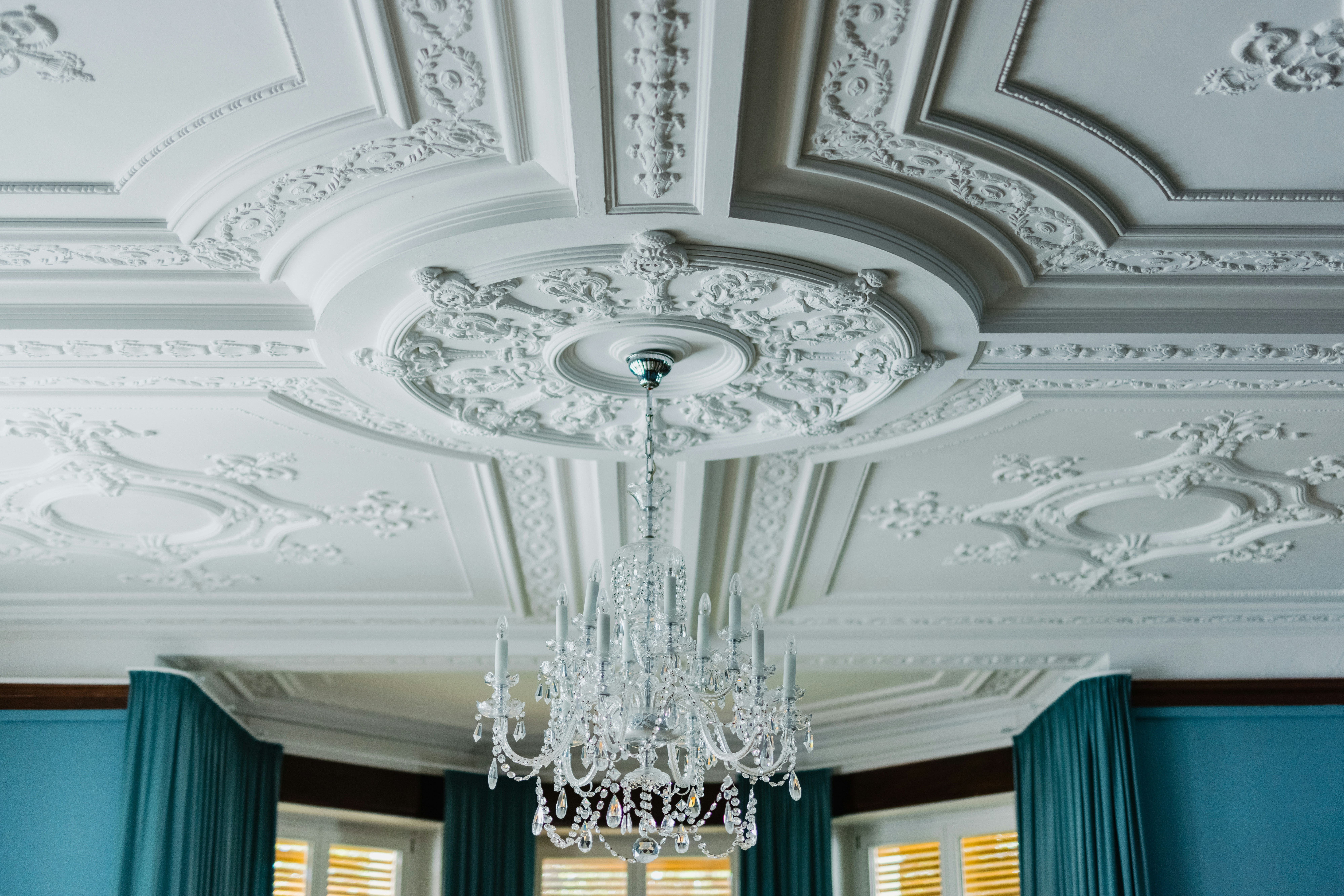 Intricately detailed white ceiling with an elegant chandelier and blue curtains framing large windows.