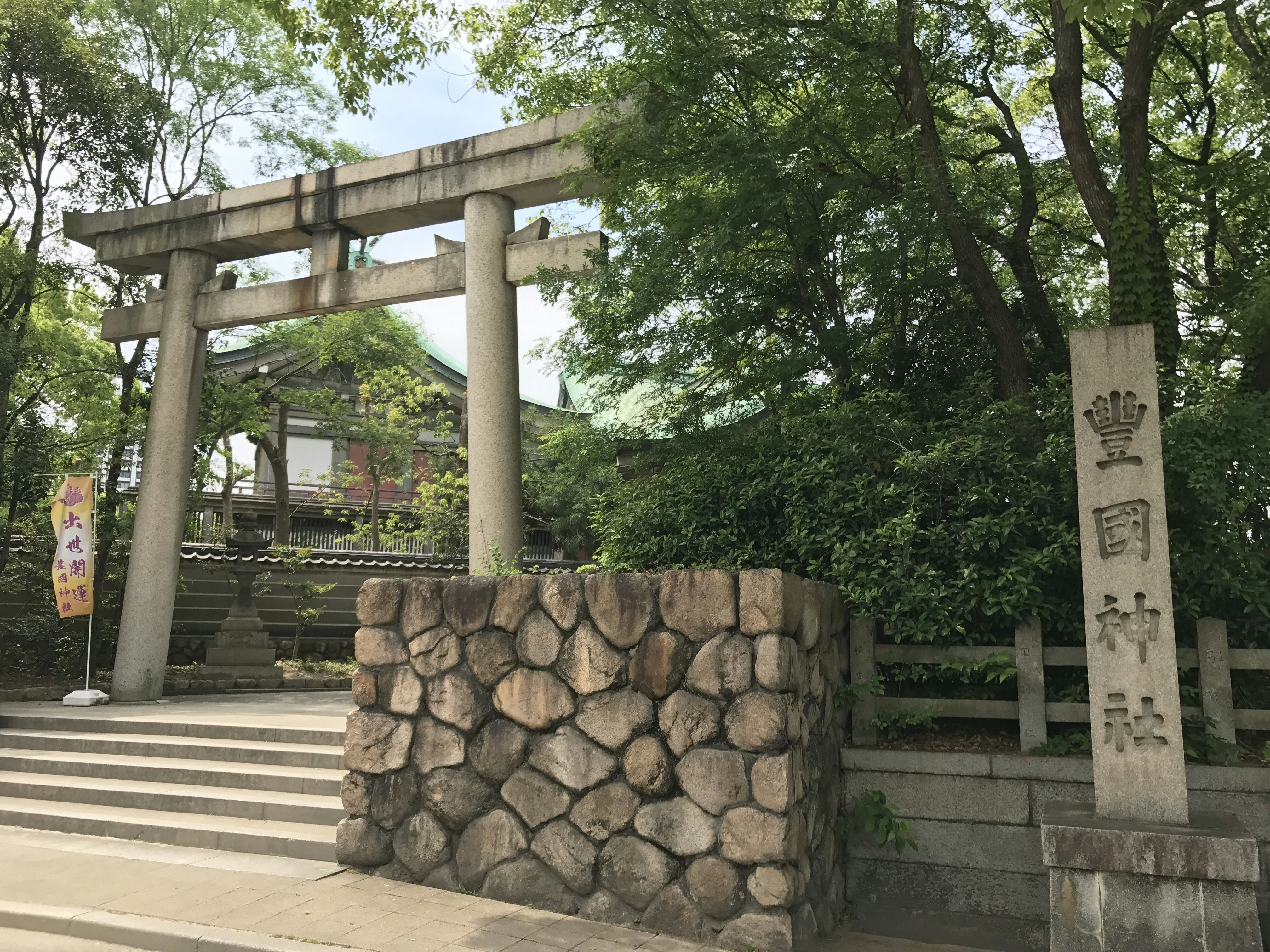Torii gate marking the entrance to a serene shrine, surrounded by lush greenery and a stone wall. The stone monument beside it adds to the cultural significance.