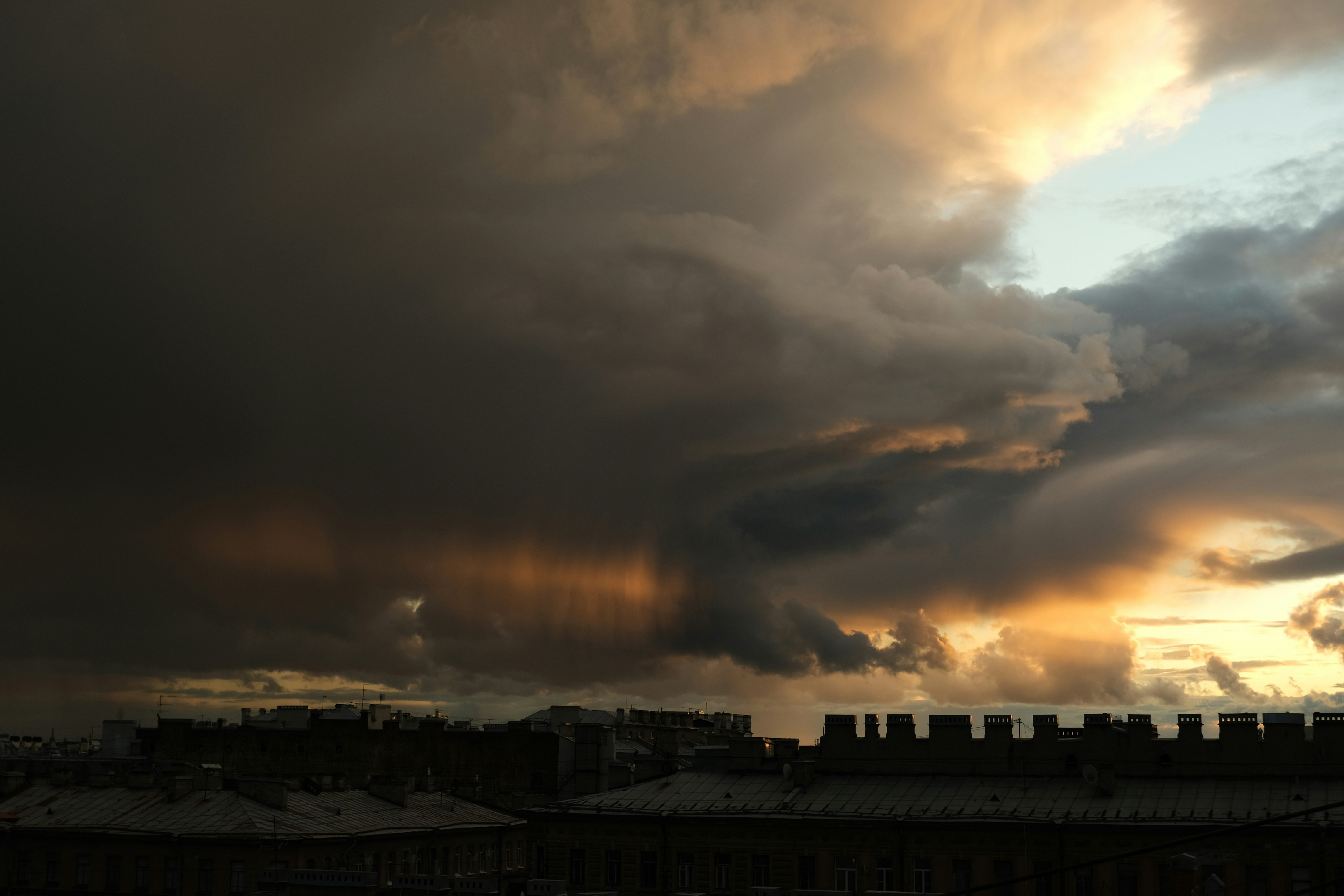 Dramatic clouds swirling above a cityscape at sunset, showcasing a blend of dark and light tones. The scene captures the dynamic beauty of changing weather.