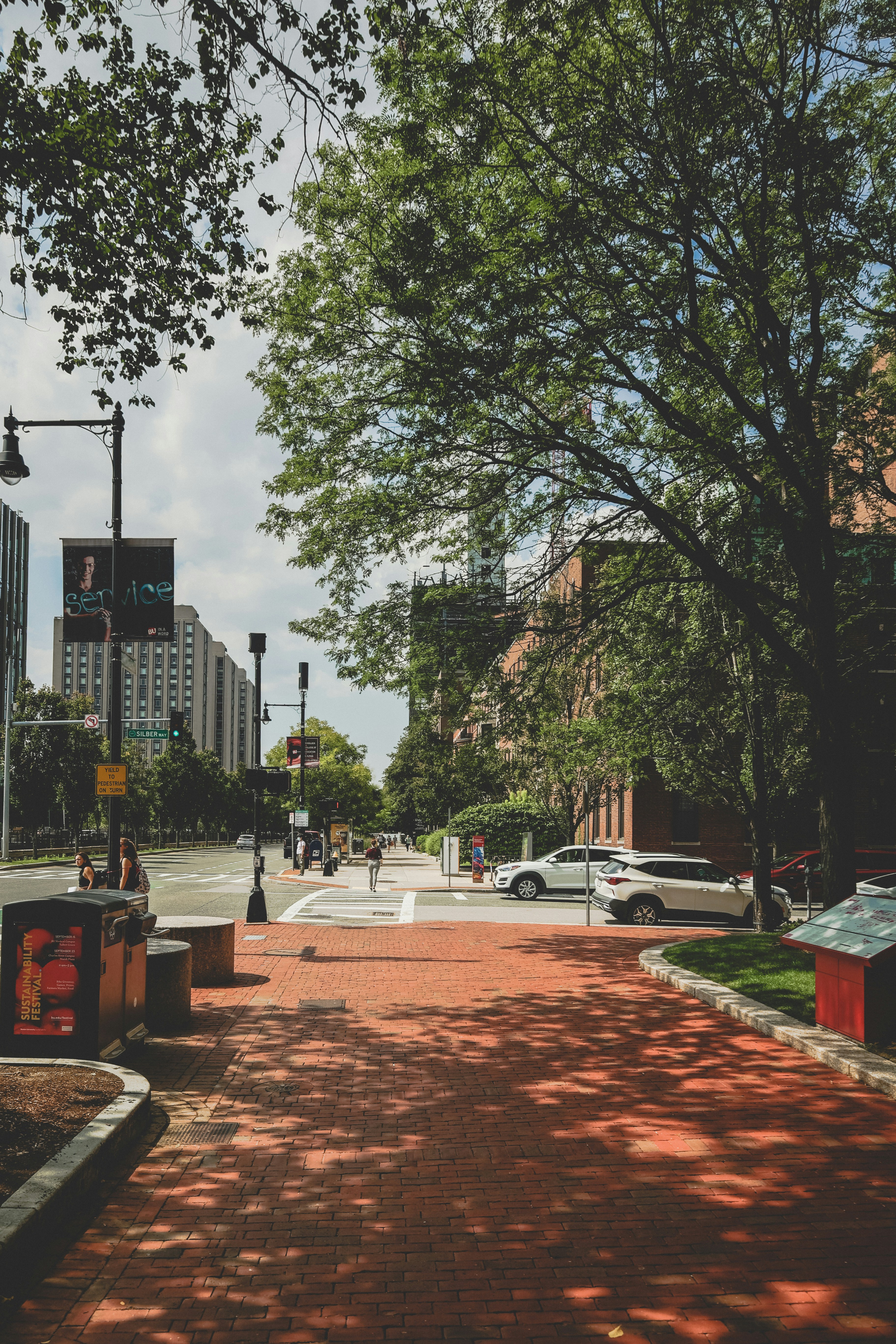 Pedestrians stroll along a brick pathway lined with trees and urban buildings, capturing the essence of city life. A vibrant billboard adds a splash of color to the scene.