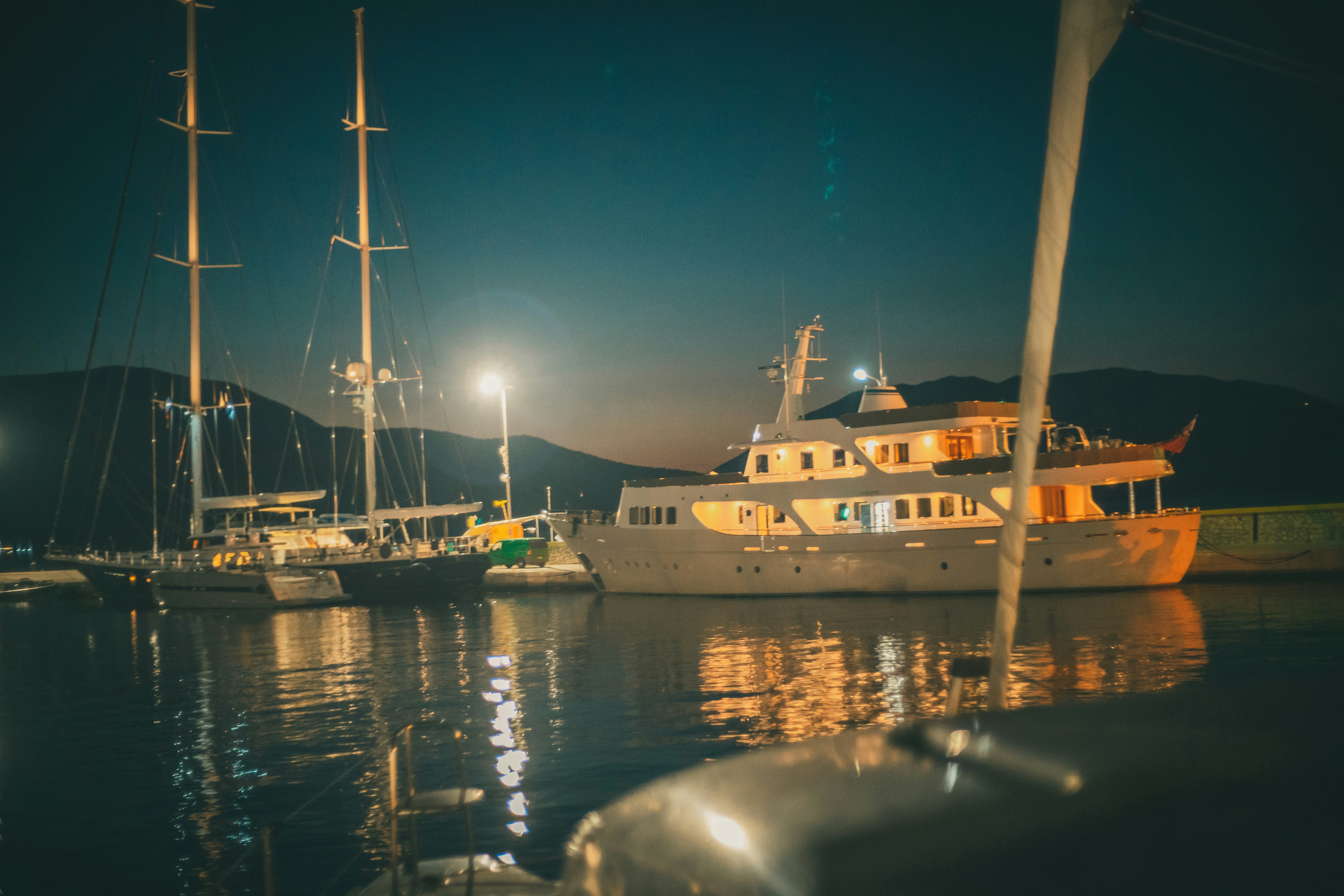 Luxury yacht illuminated in harbor at twilight, surrounded by sailboats and gentle water reflections.