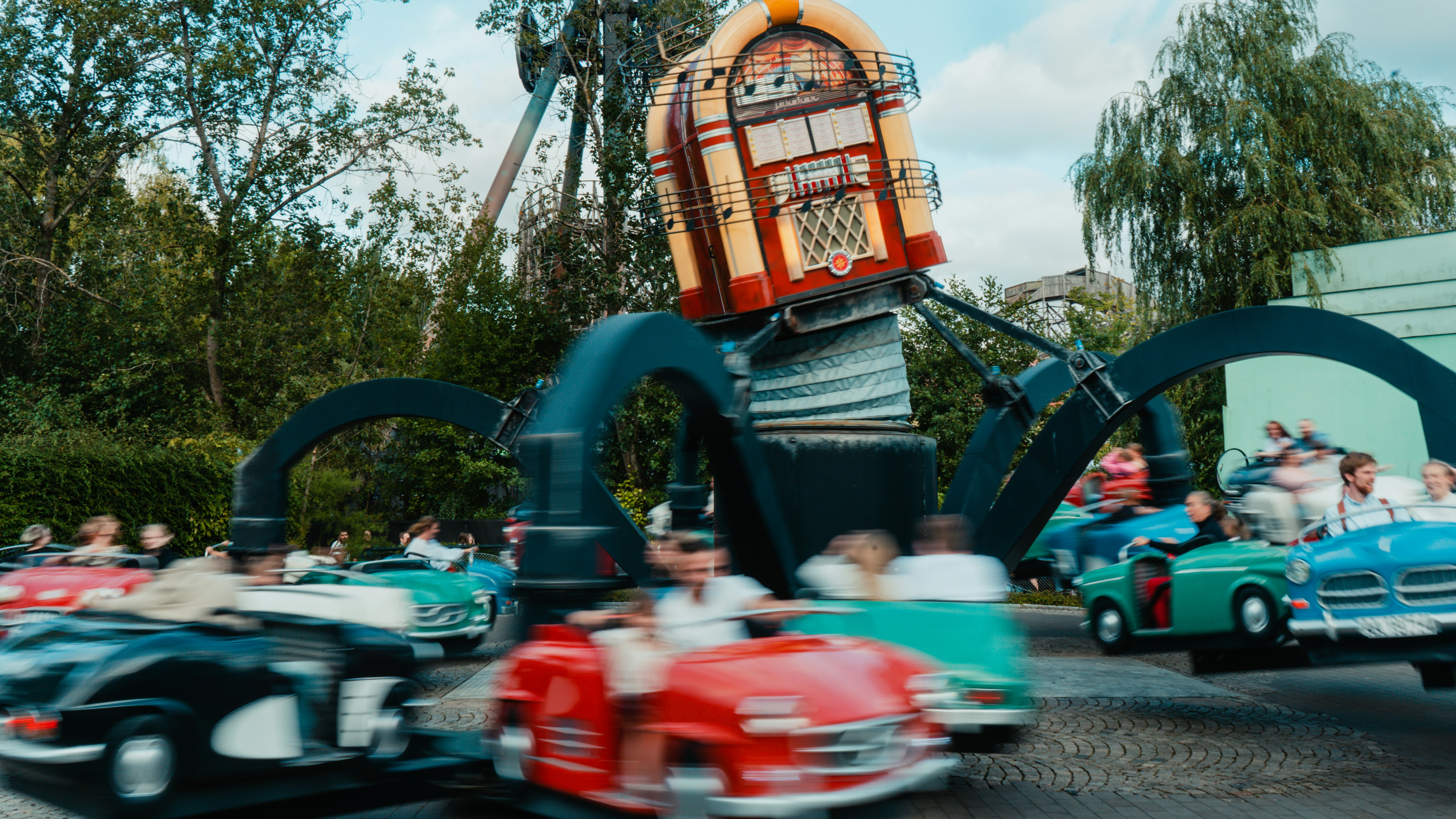 A vibrant amusement park ride featuring colorful vintage cars zooming around a central jukebox structure, evoking a sense of childhood joy and excitement.