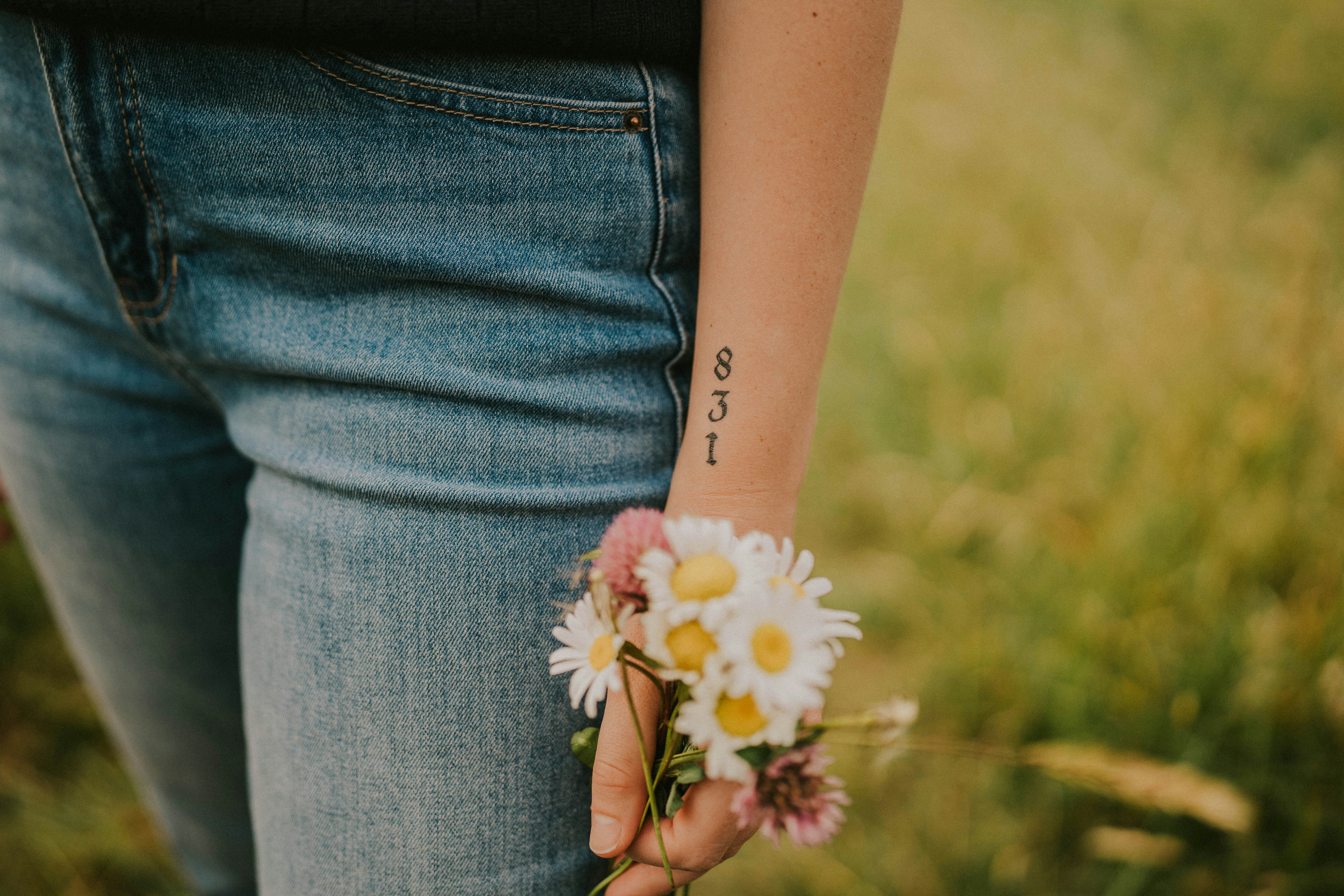A hand adorned with a floral bouquet gently rests against a denim-clad thigh, showcasing a meaningful tattoo. The background features a soft, blurred green landscape.