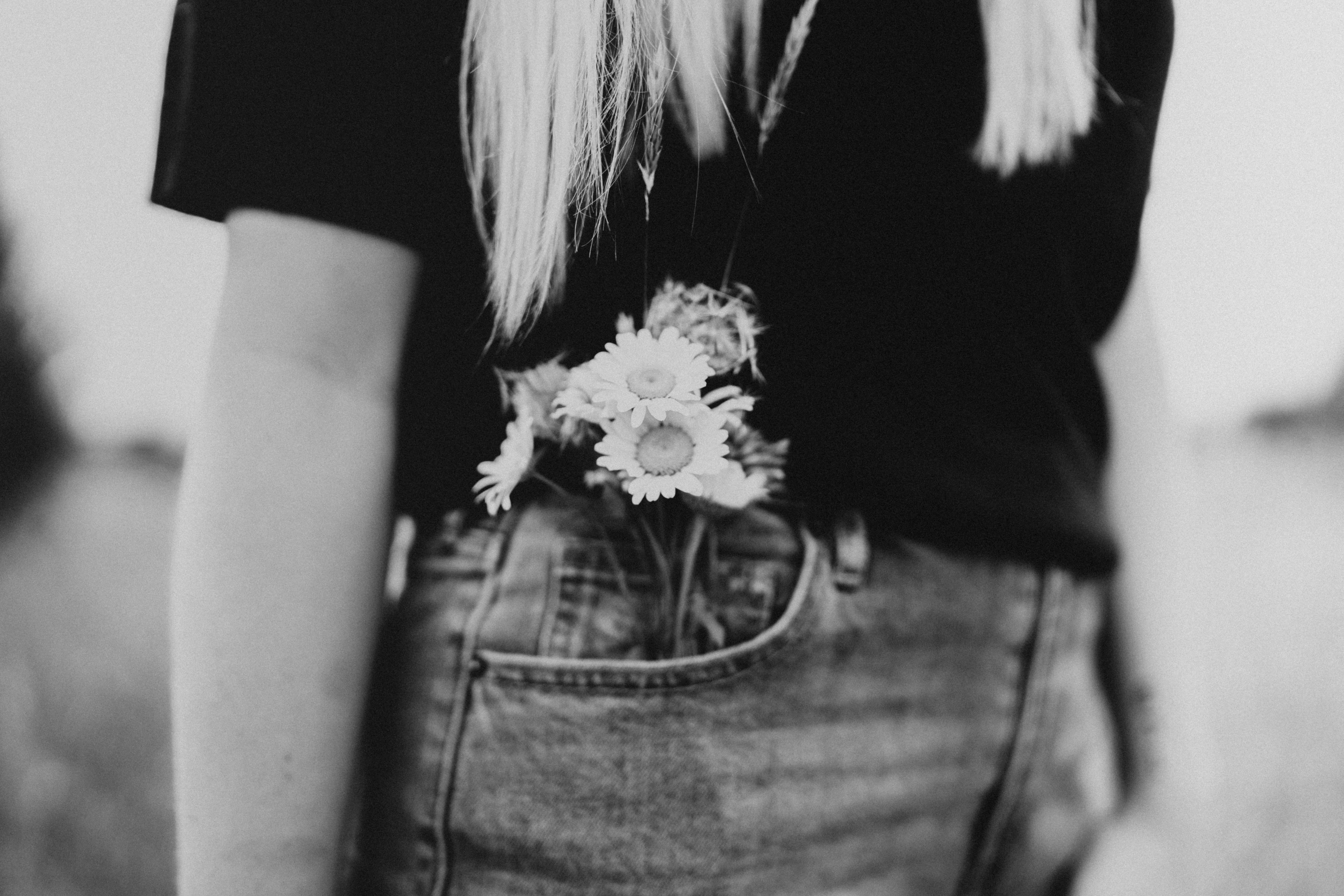 A close-up of a person holding flowers in the pocket of their jeans, set against a softly blurred natural background.