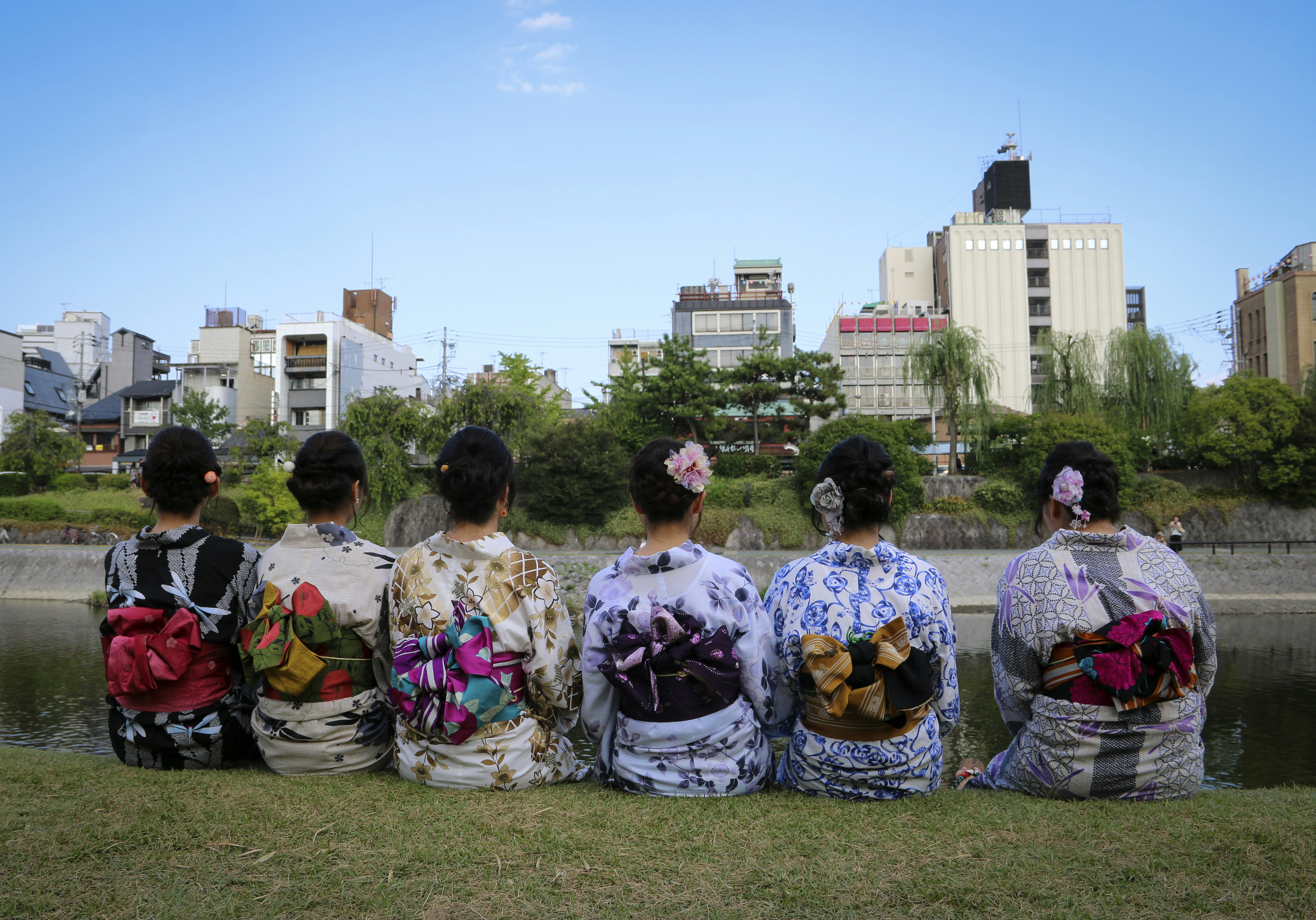 Women in colorful kimonos seated by a river, showcasing intricate obi designs against an urban backdrop. A moment of cultural reflection and connection.