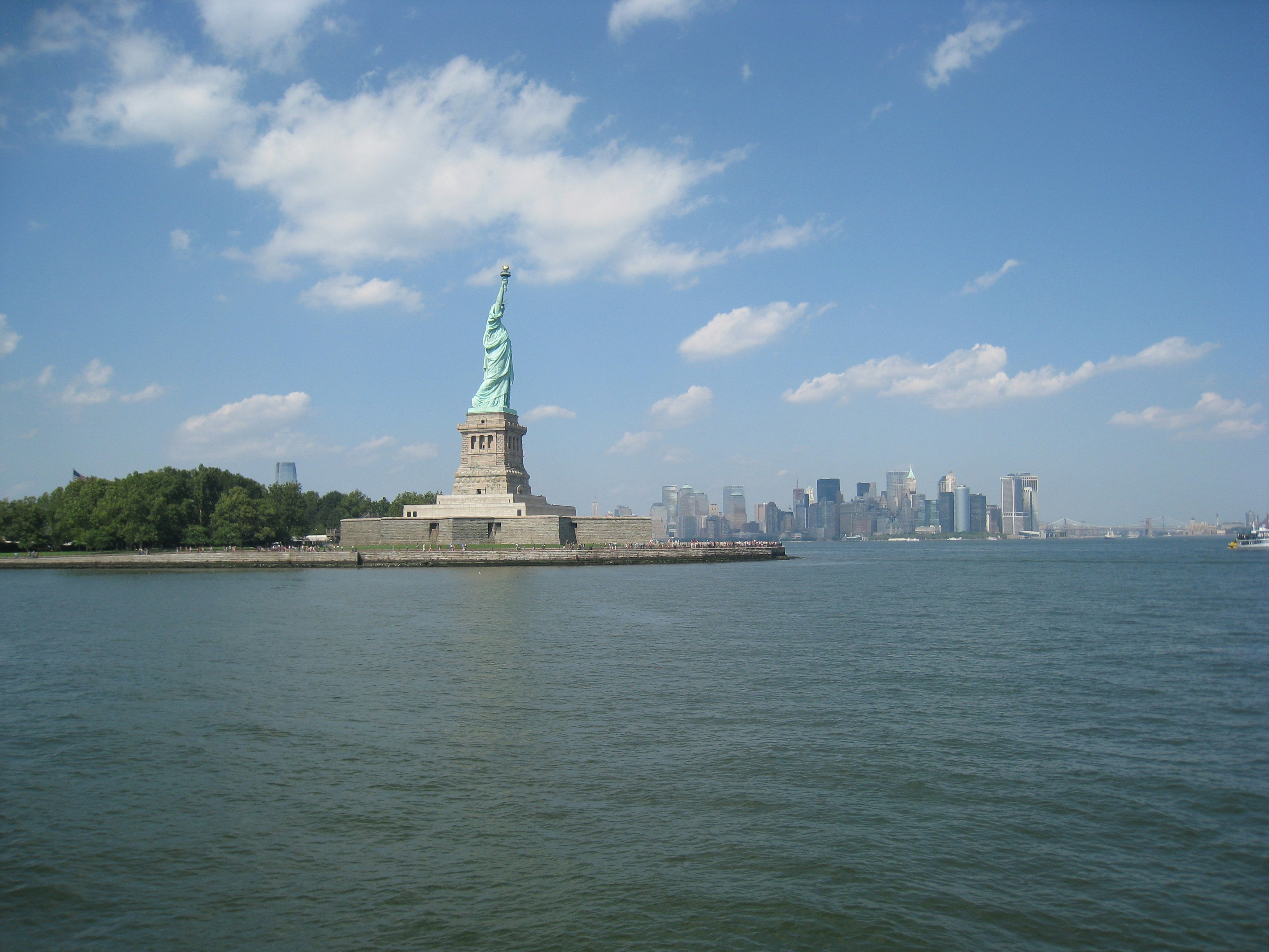 Statue of Liberty stands on Liberty Island beside calm harbor water, with a distant Manhattan skyline under a clear blue sky.