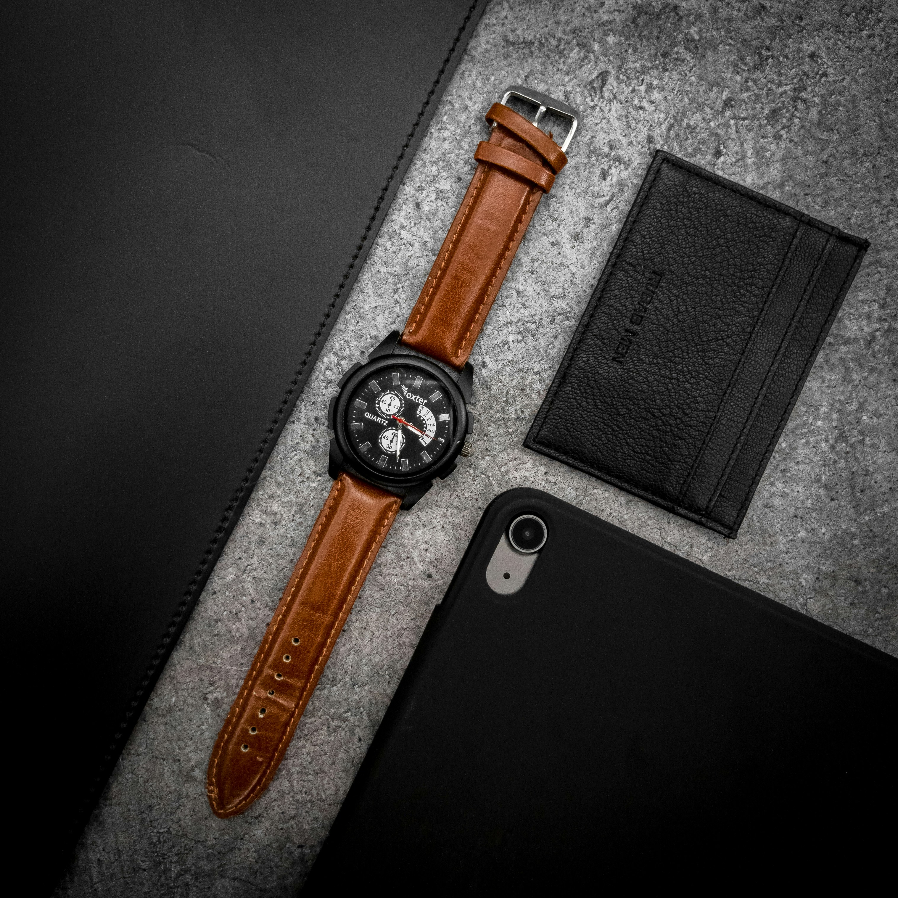 Close-up still life of a brown-leather watch laid beside a black wallet and a smartphone on a textured gray surface.