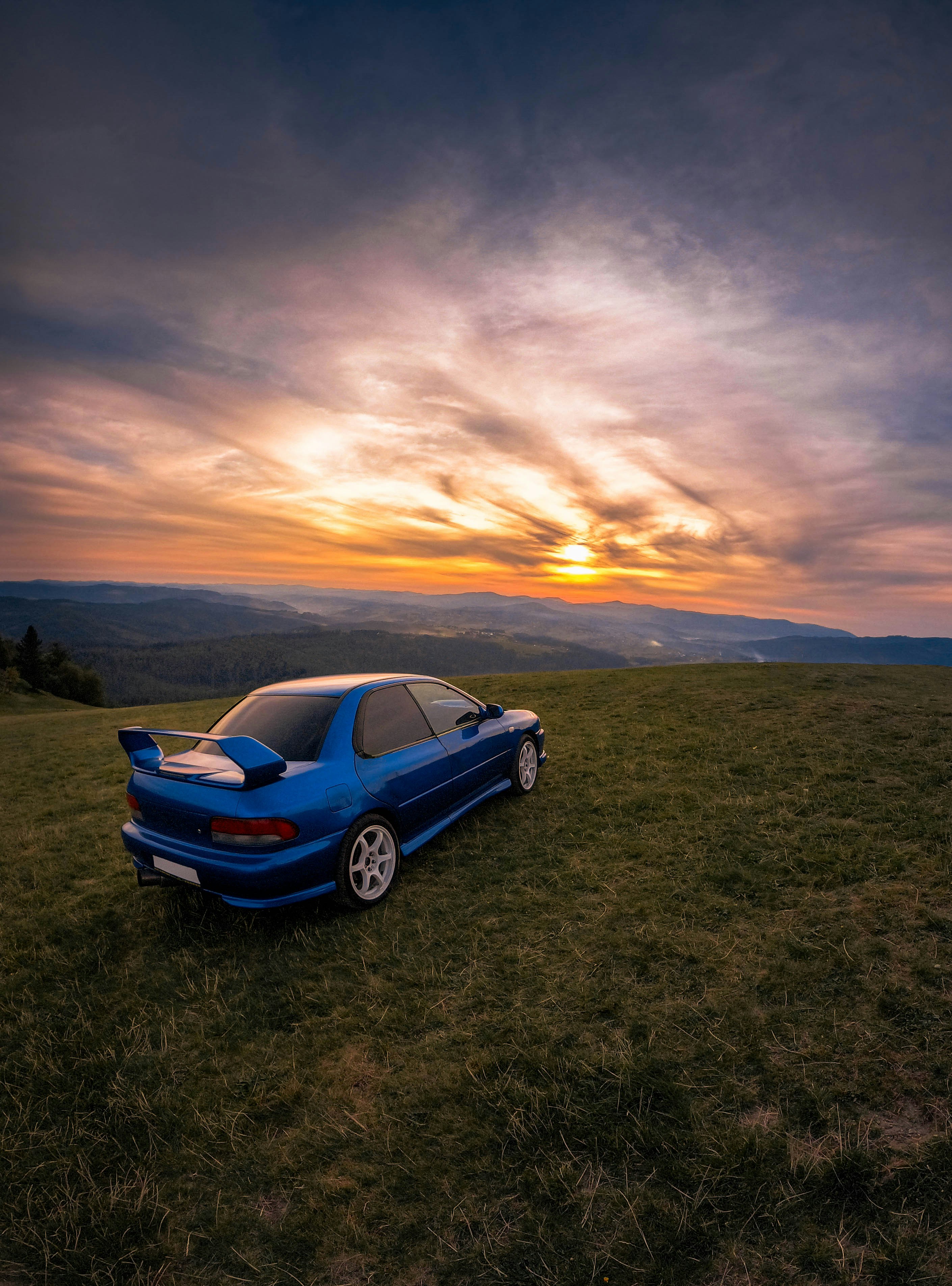 A vibrant blue sports car parked on a grassy hill, overlooking a vast landscape at sunset, with dramatic clouds filling the sky.