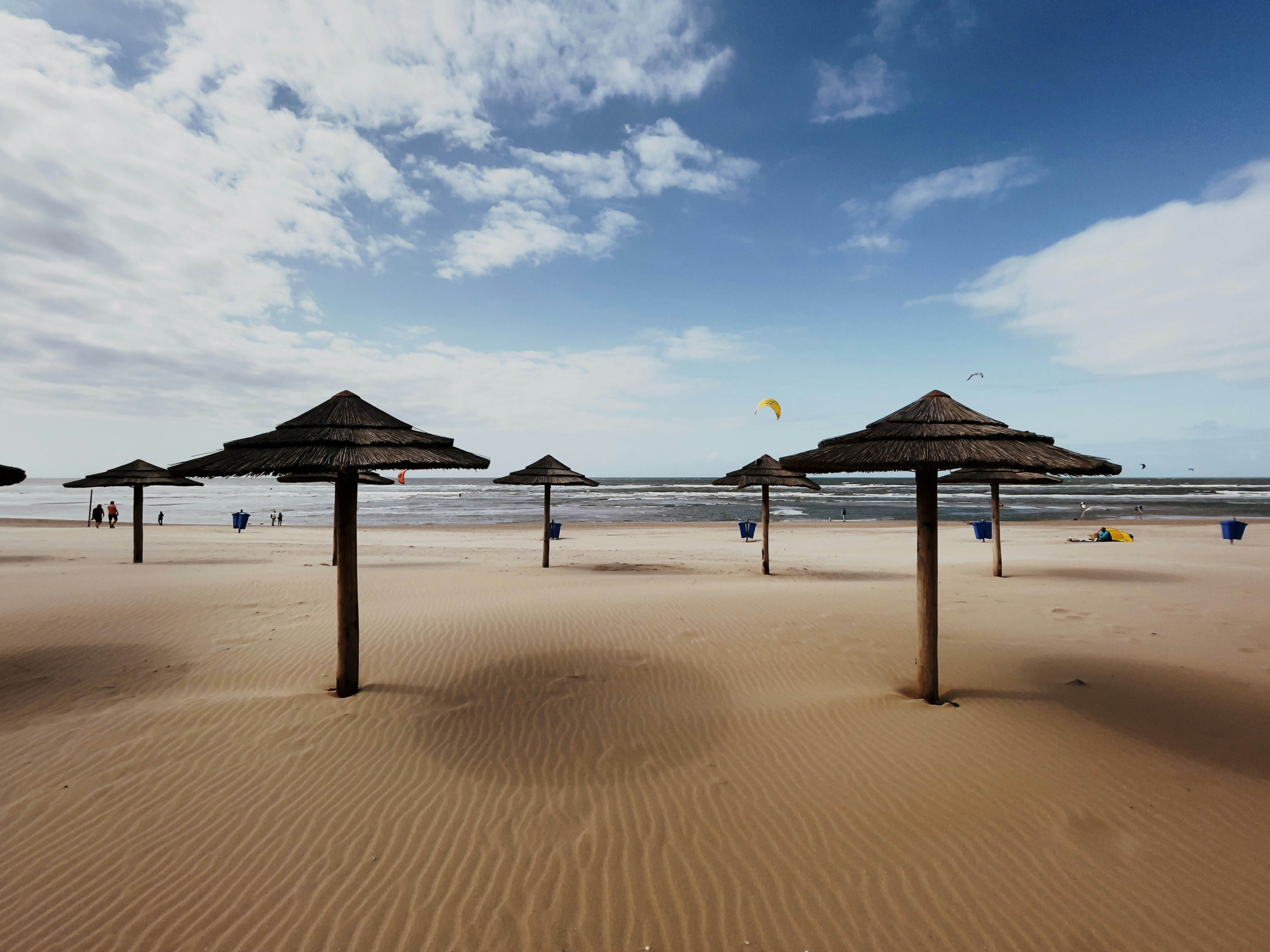 Beach umbrellas stand tall on golden sands, framing a tranquil seascape under a partly cloudy sky. Kitesurfers dot the horizon, adding a sense of adventure.