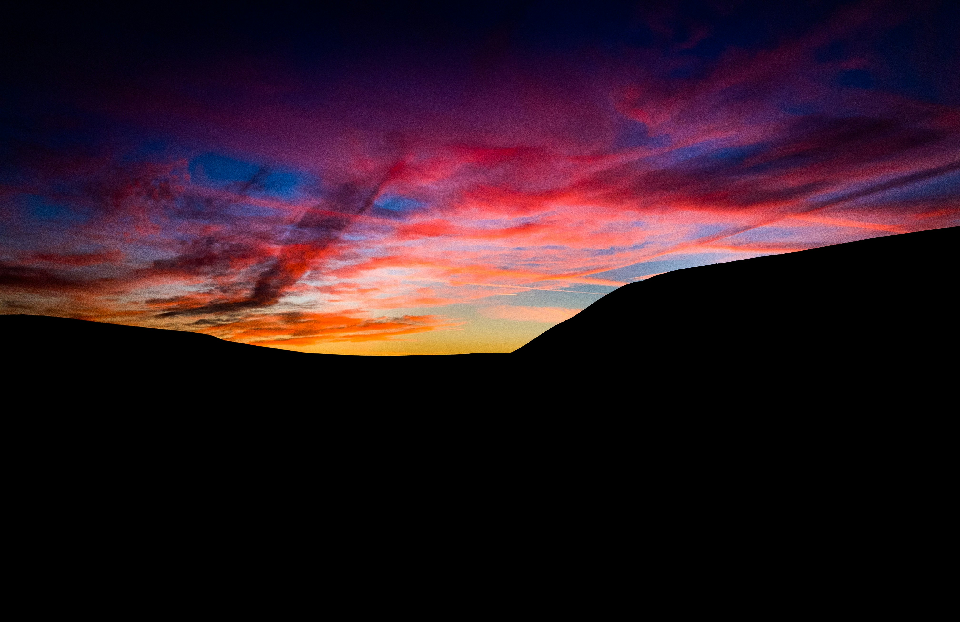 Vibrant sunset hues blend seamlessly across the horizon, silhouetting rolling hills against a dramatic sky.