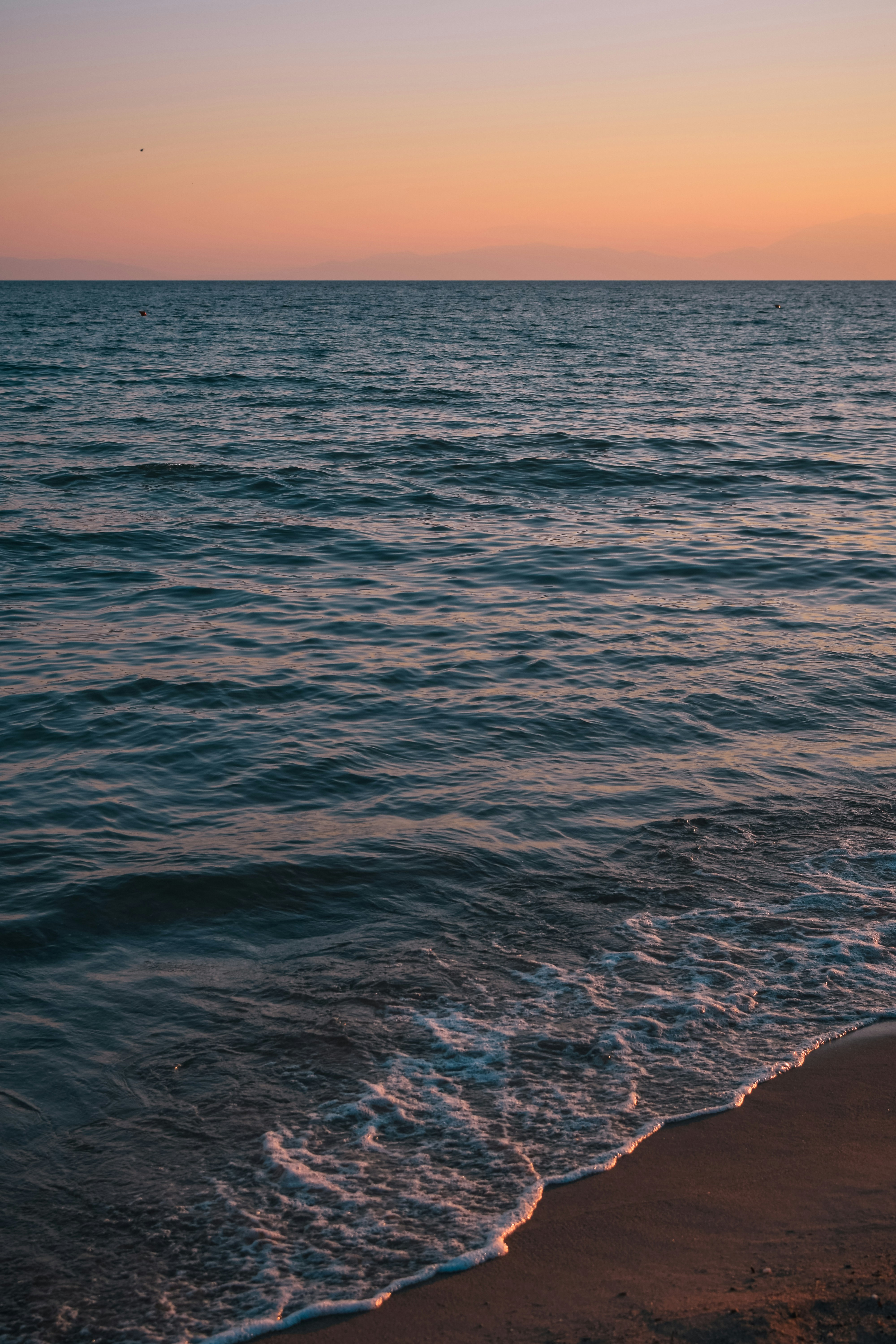 Gentle waves lap against the sandy shoreline under a warm, orange sunset sky.