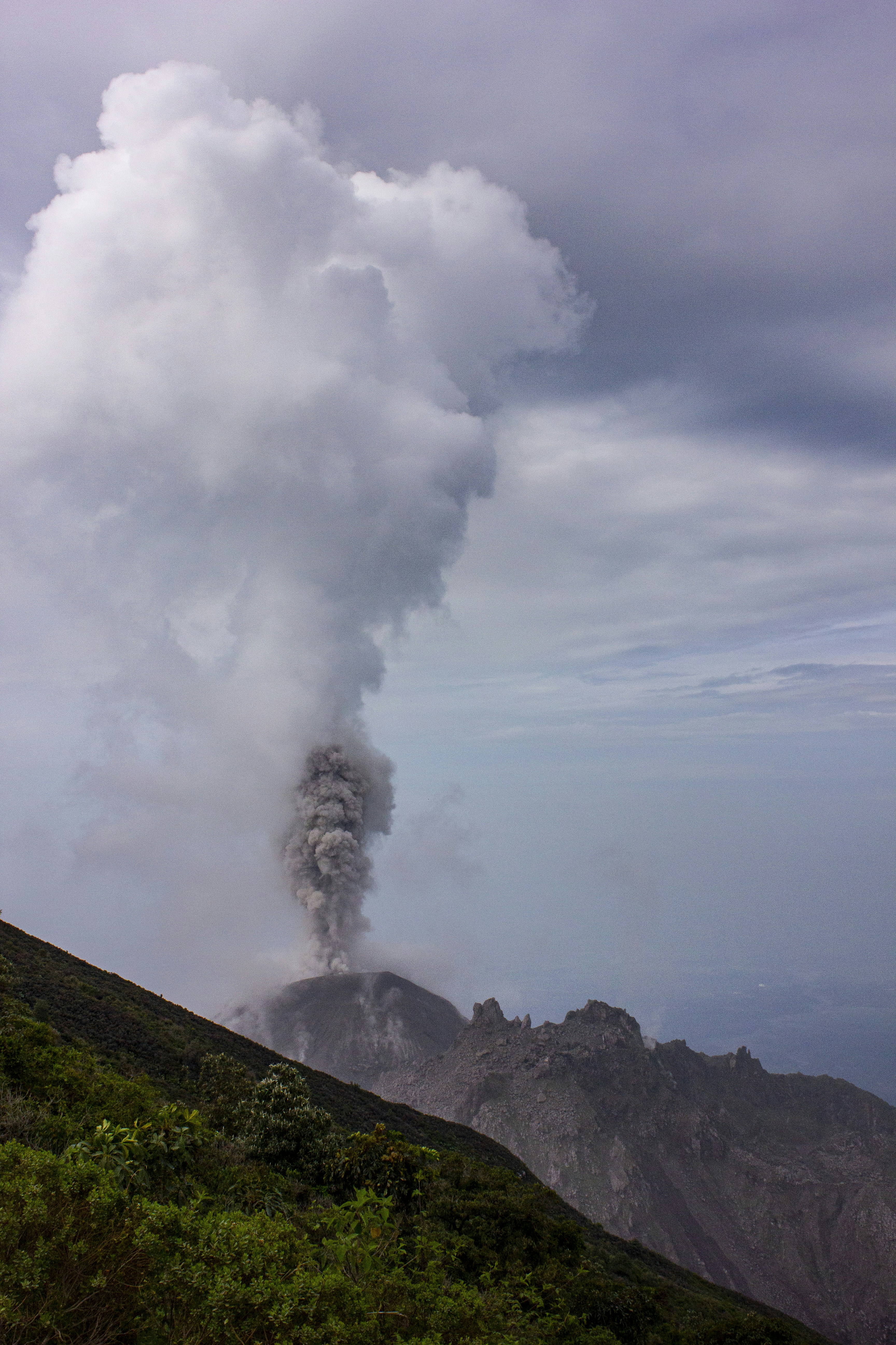 Volcanic eruption spewing ash and smoke against a dramatic sky, framed by rugged mountain terrain.