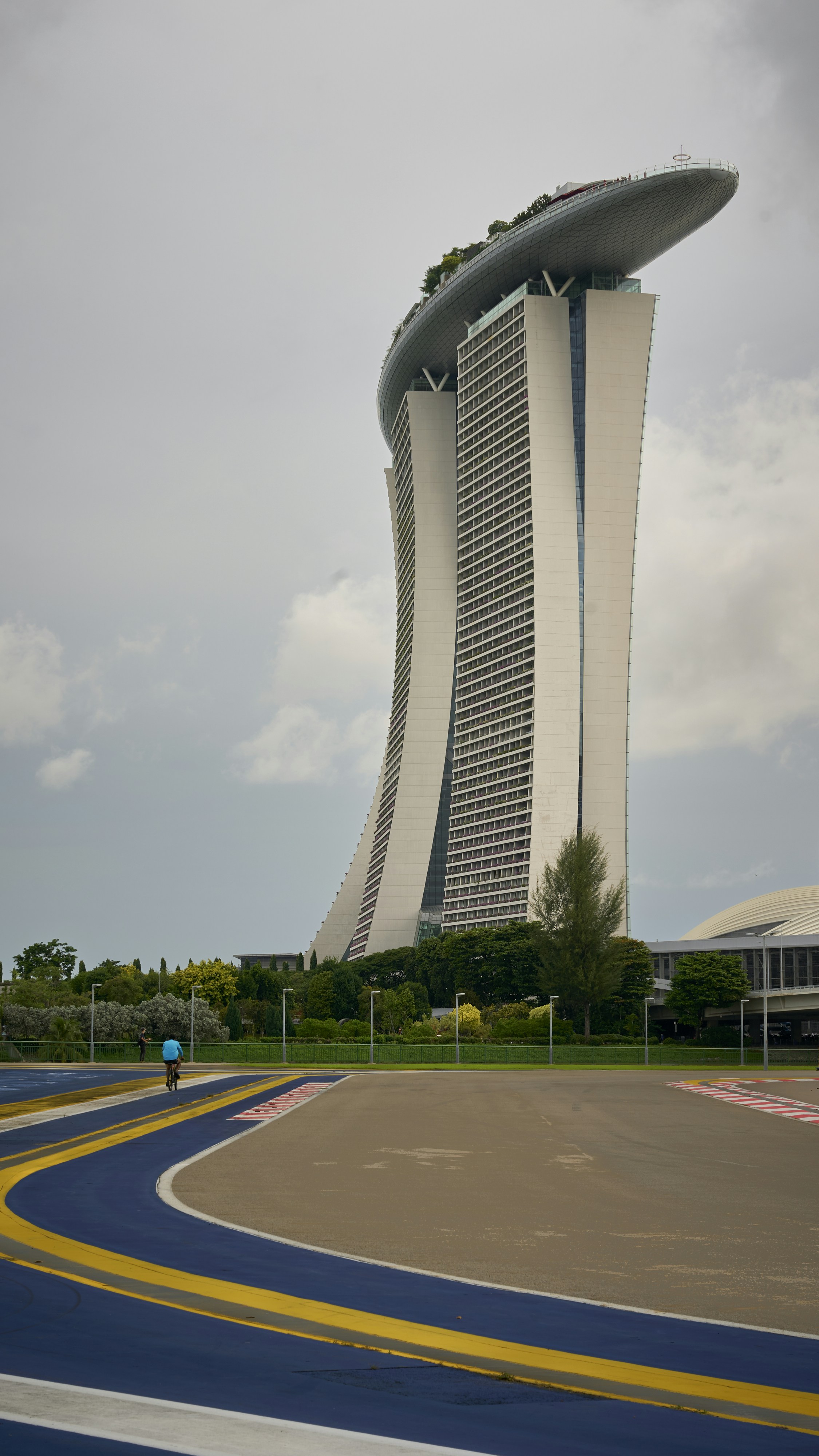 A striking modern skyscraper with a unique curved design, set against a cloudy sky and surrounded by lush greenery. A lone figure walks along a winding path in the foreground.