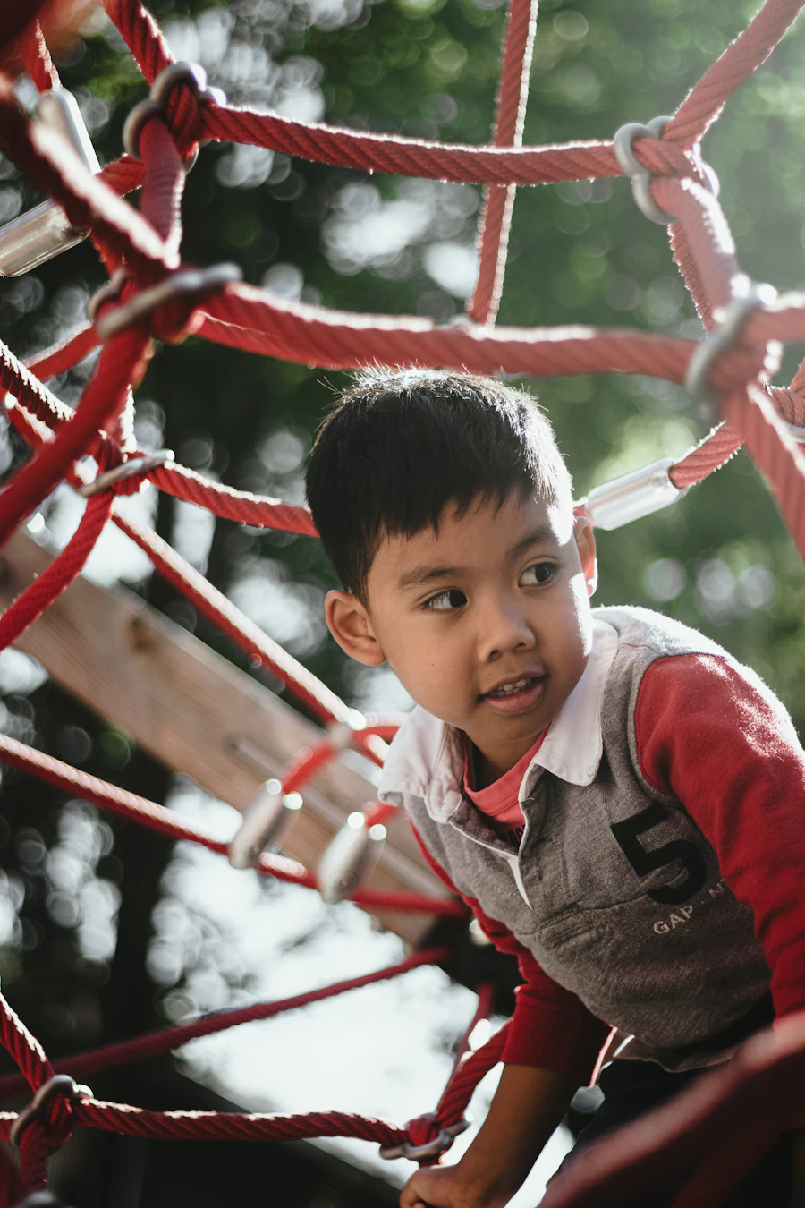 A young boy explores a red rope climbing net in a sunlit playground, his gaze focused on his next move.
