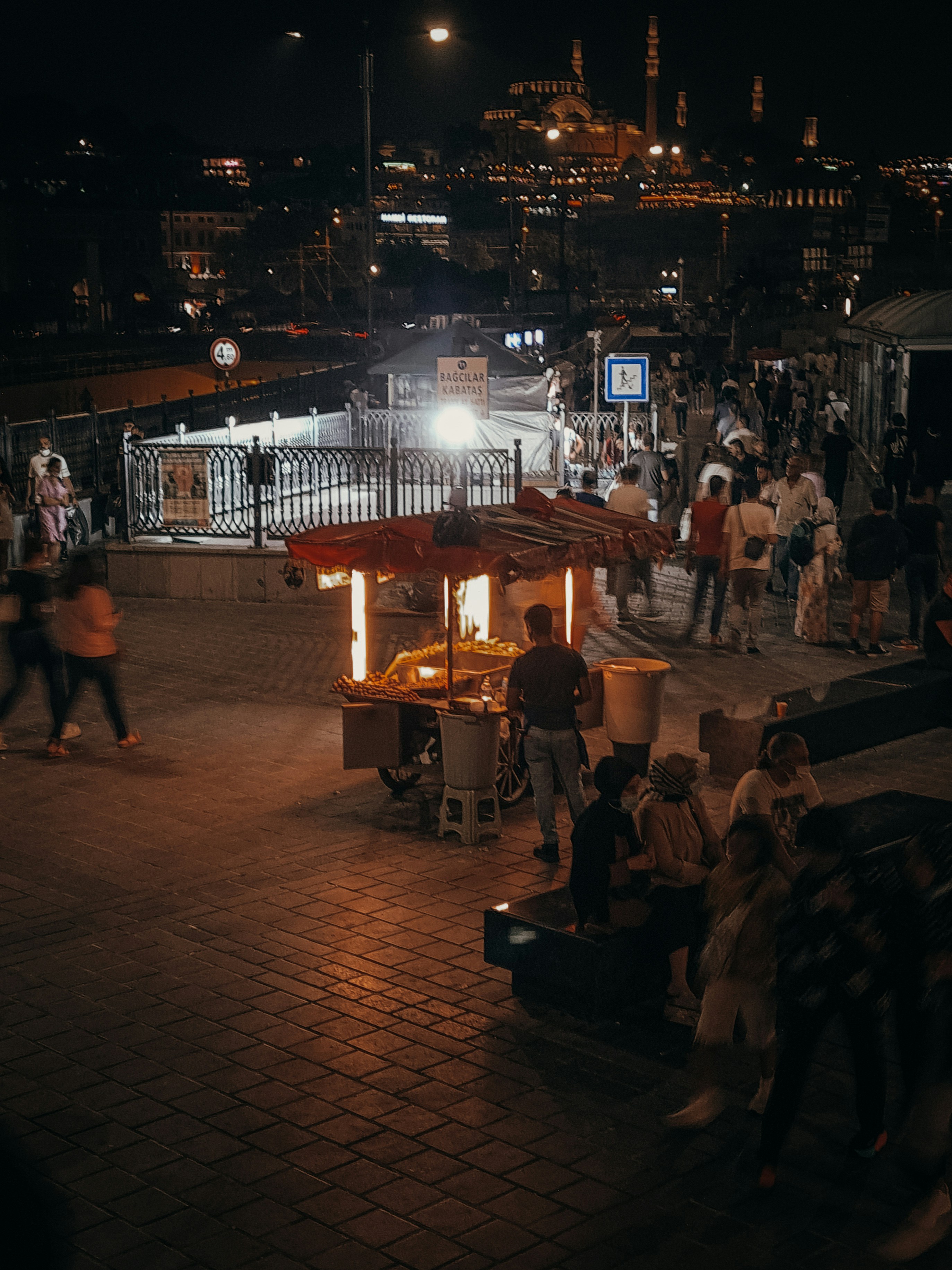 Vibrant street scene featuring a food cart illuminated by warm lights, surrounded by bustling pedestrians at night. The backdrop includes historical architecture, adding depth to the urban atmosphere.