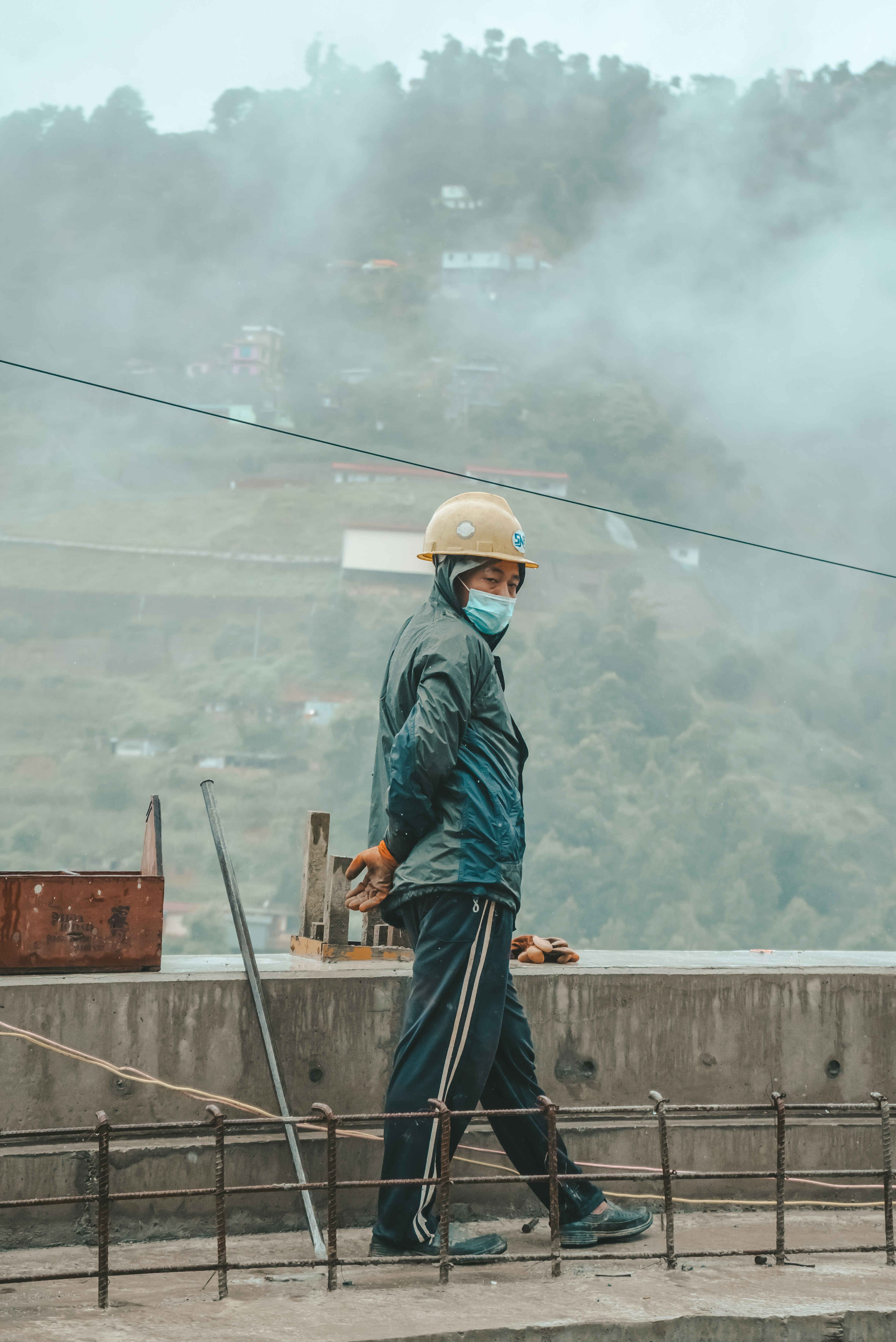 Construction worker in a blue jacket and hard hat navigating a foggy building site with distant greenery in the background.