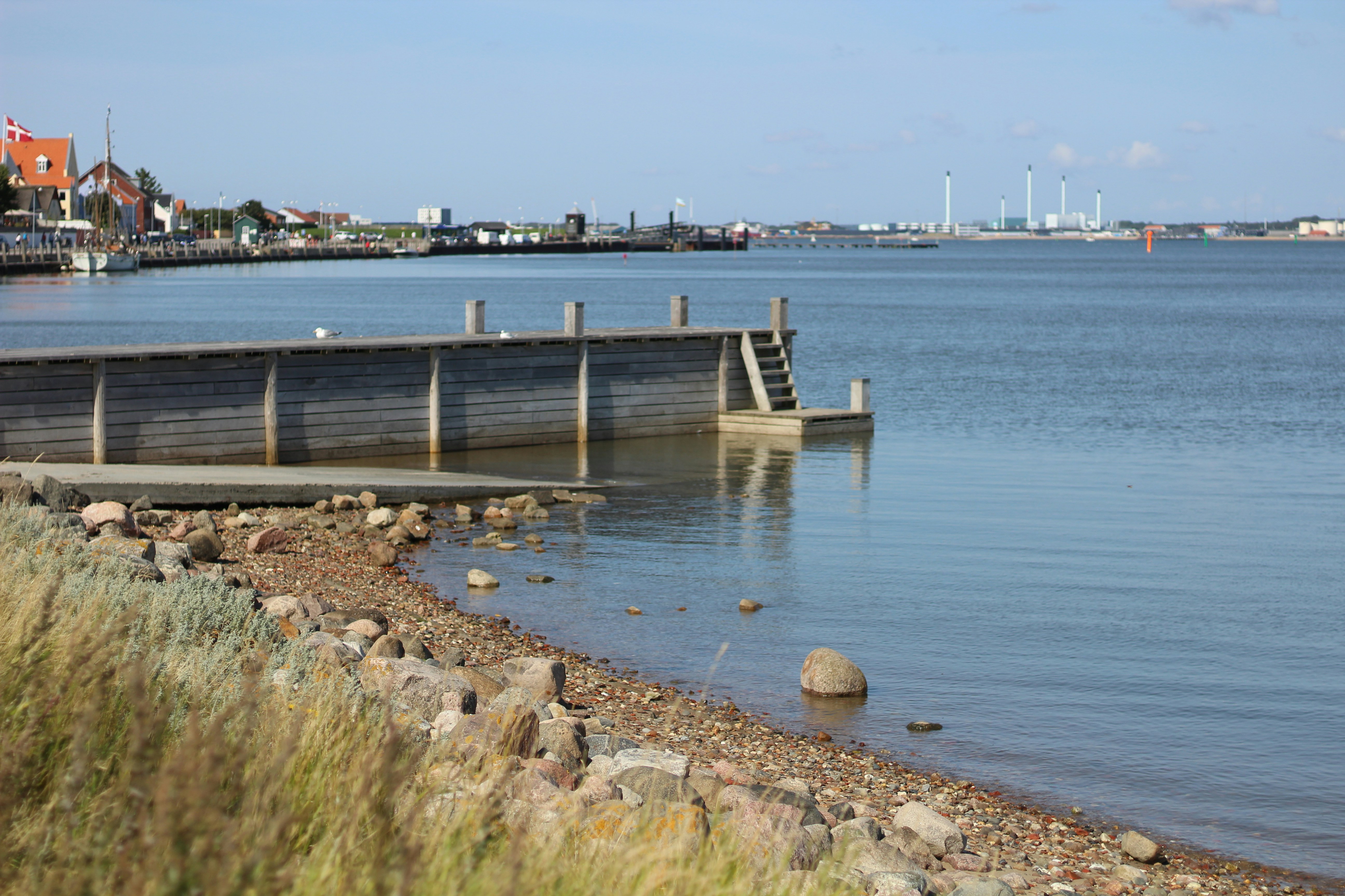 Wooden dock extending into calm waters, framed by rocky shoreline and distant wind turbines. Lush grass adds a natural touch.
