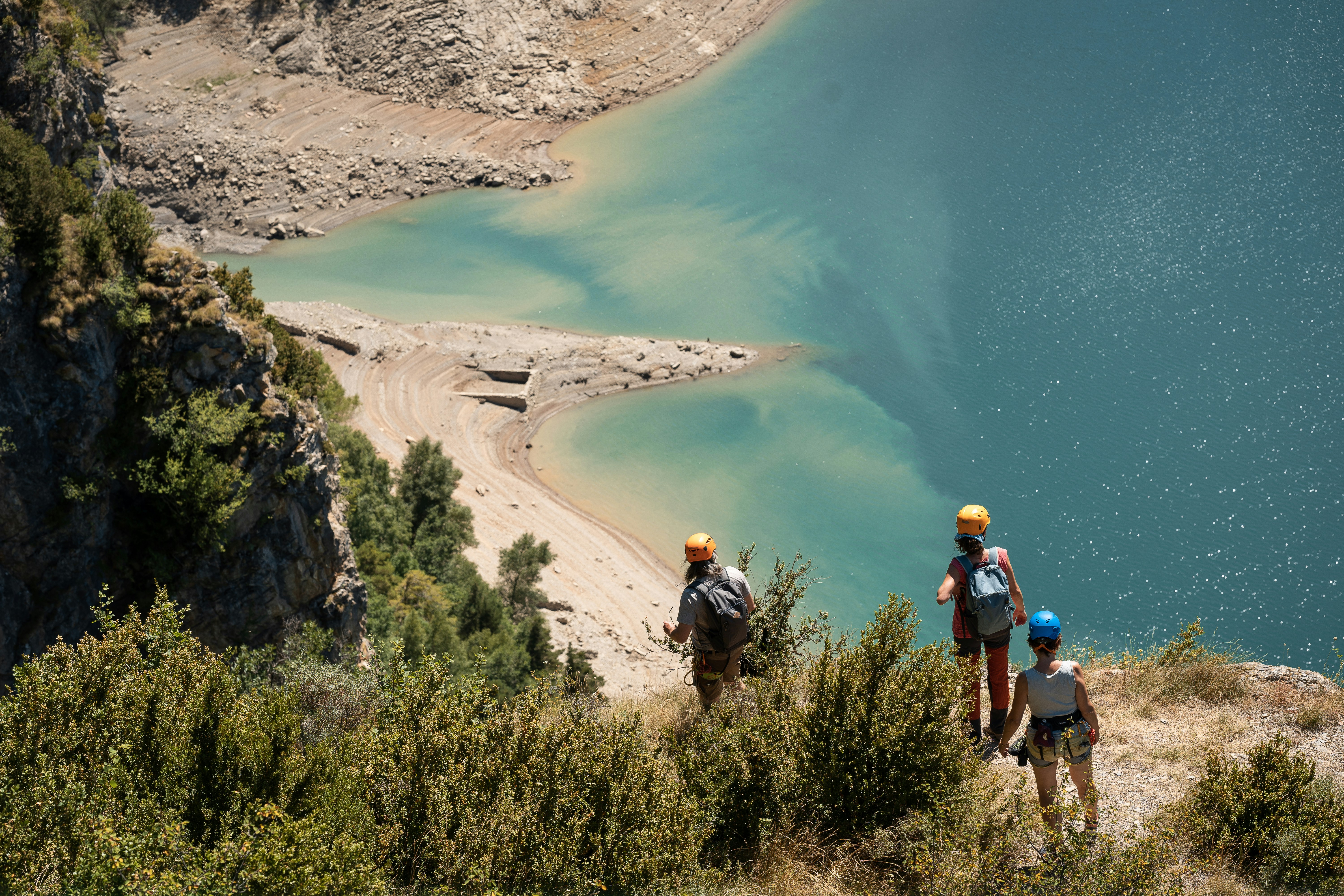 Hikers with helmets stand on a mountain edge overlooking a vibrant turquoise lake.