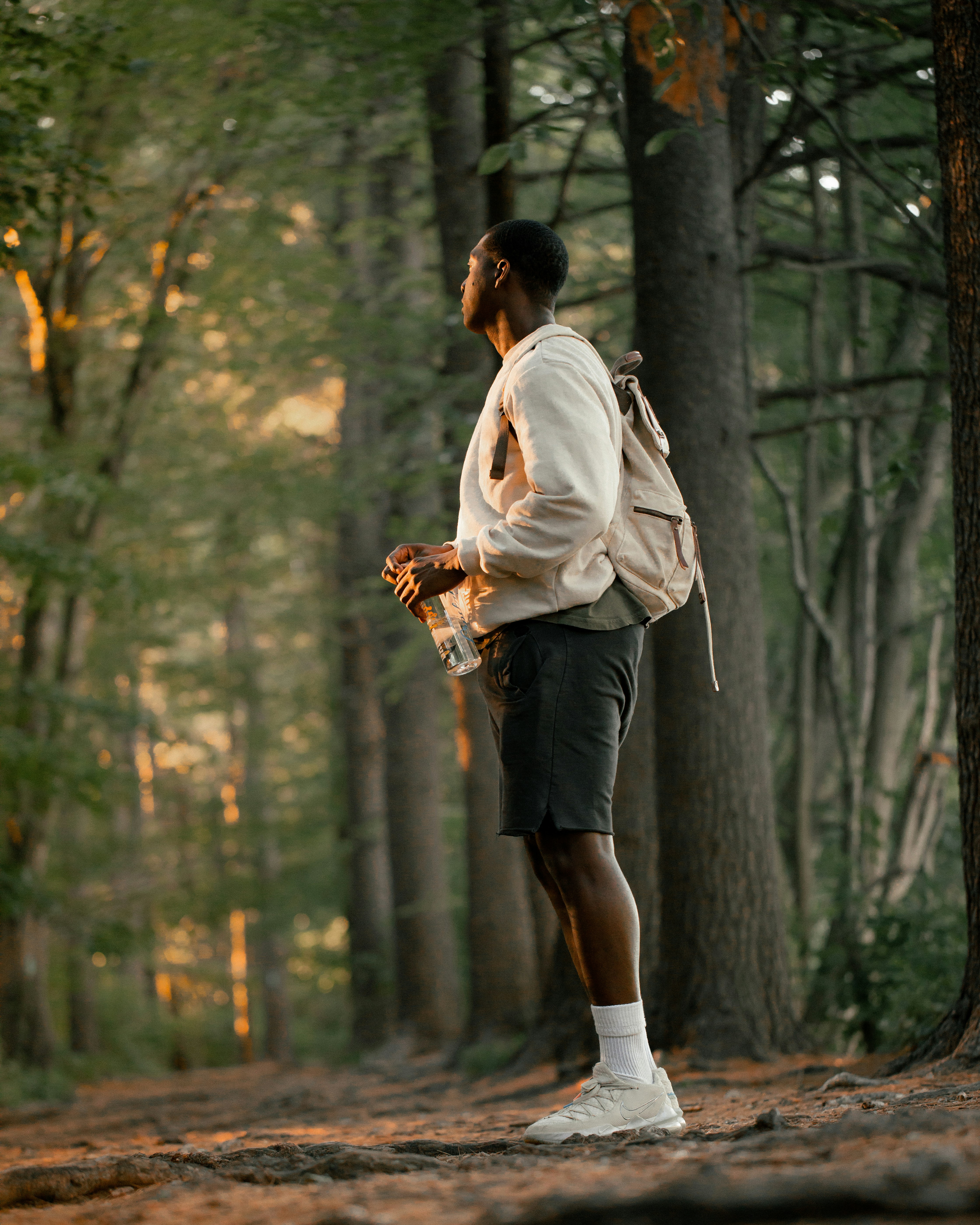 Man standing in a sunlit forest, holding a water bottle and gazing thoughtfully into the distance.