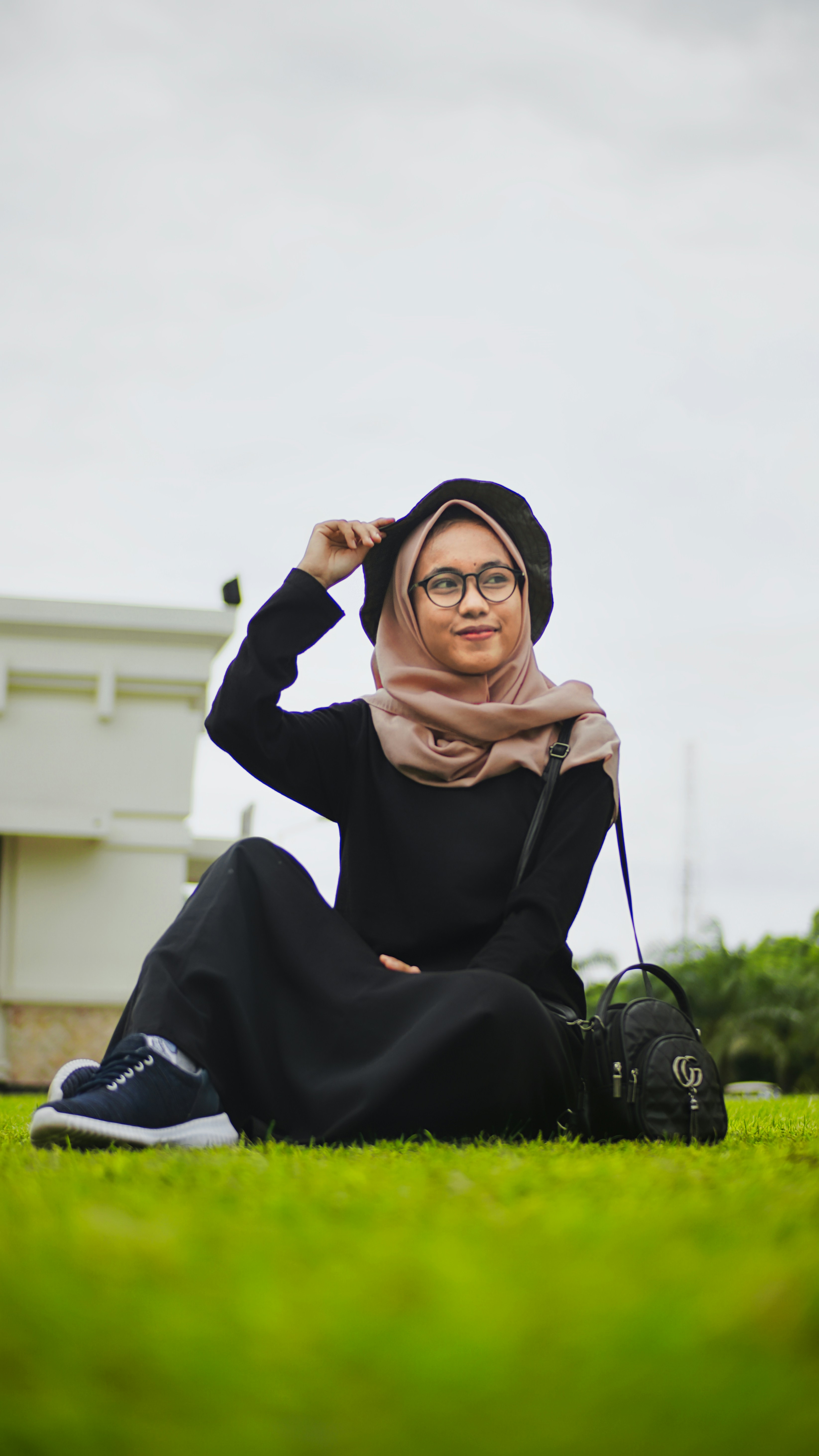 Woman in a black outfit with a brown hijab sitting on grass, touching the brim of her hat.