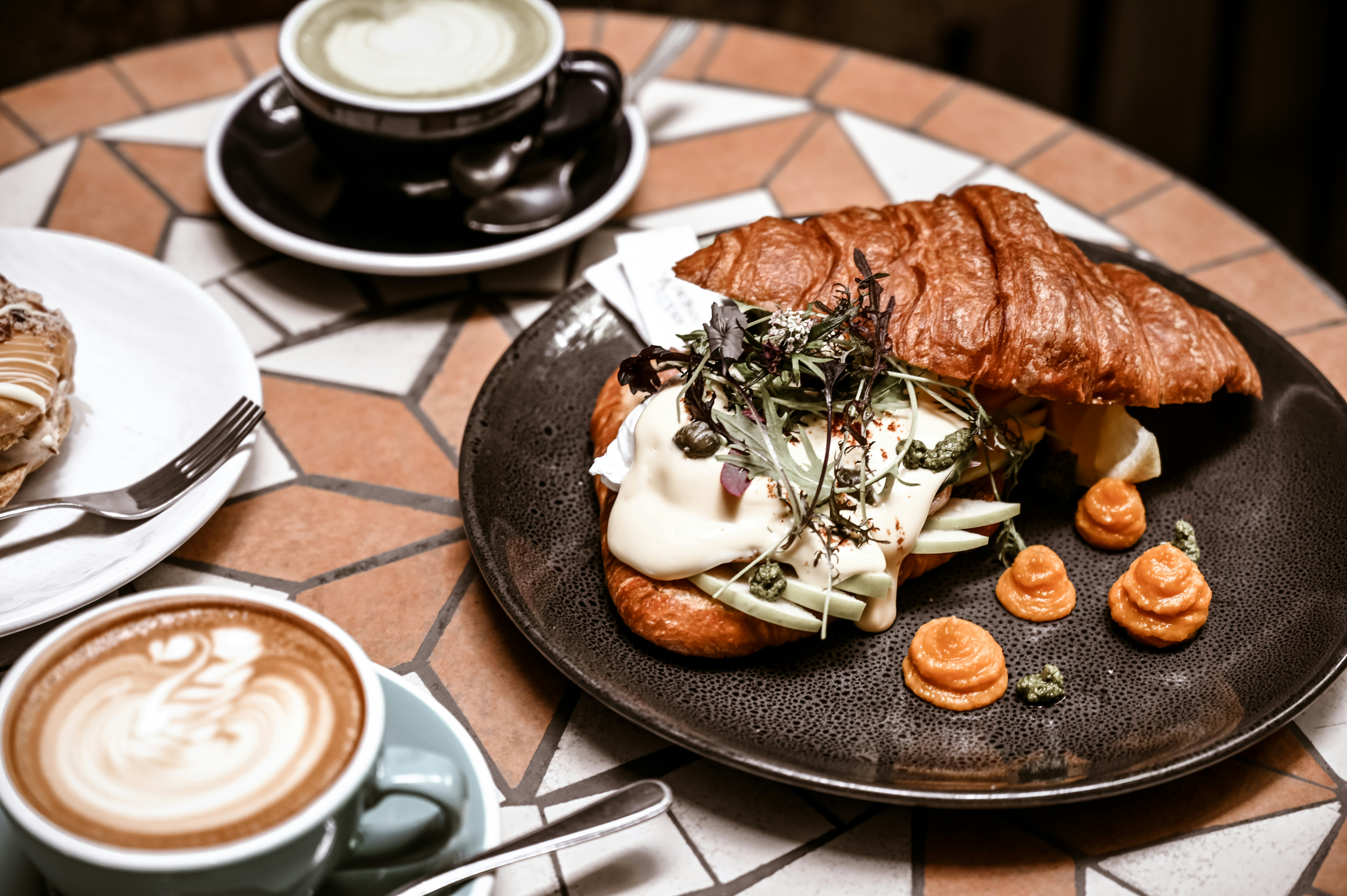 Herb-topped croissant sandwich on a dark plate amid a tiled cafe scene