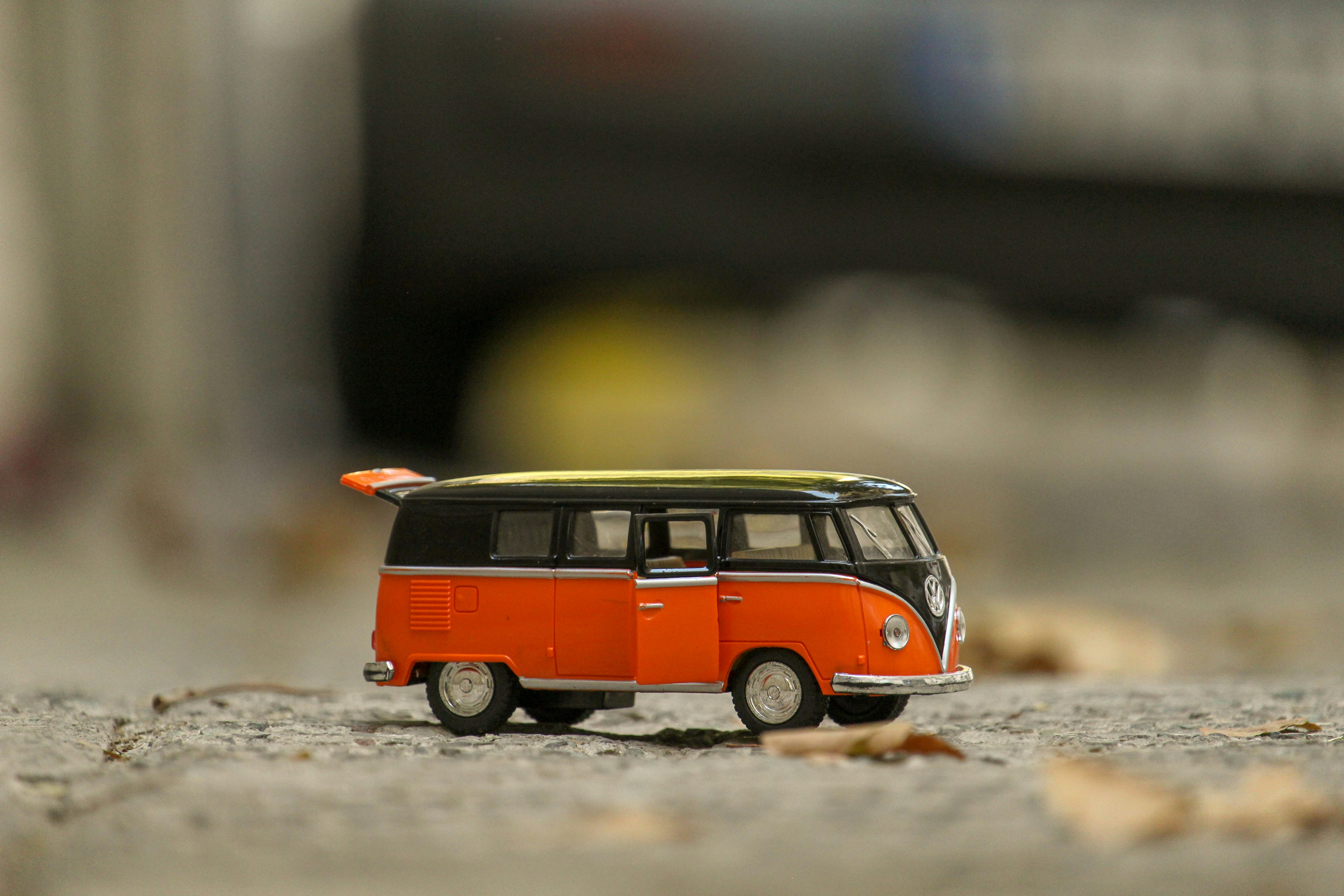 A vibrant orange and black toy van rests on a textured surface scattered with autumn leaves. The scene evokes a sense of nostalgia and adventure.
